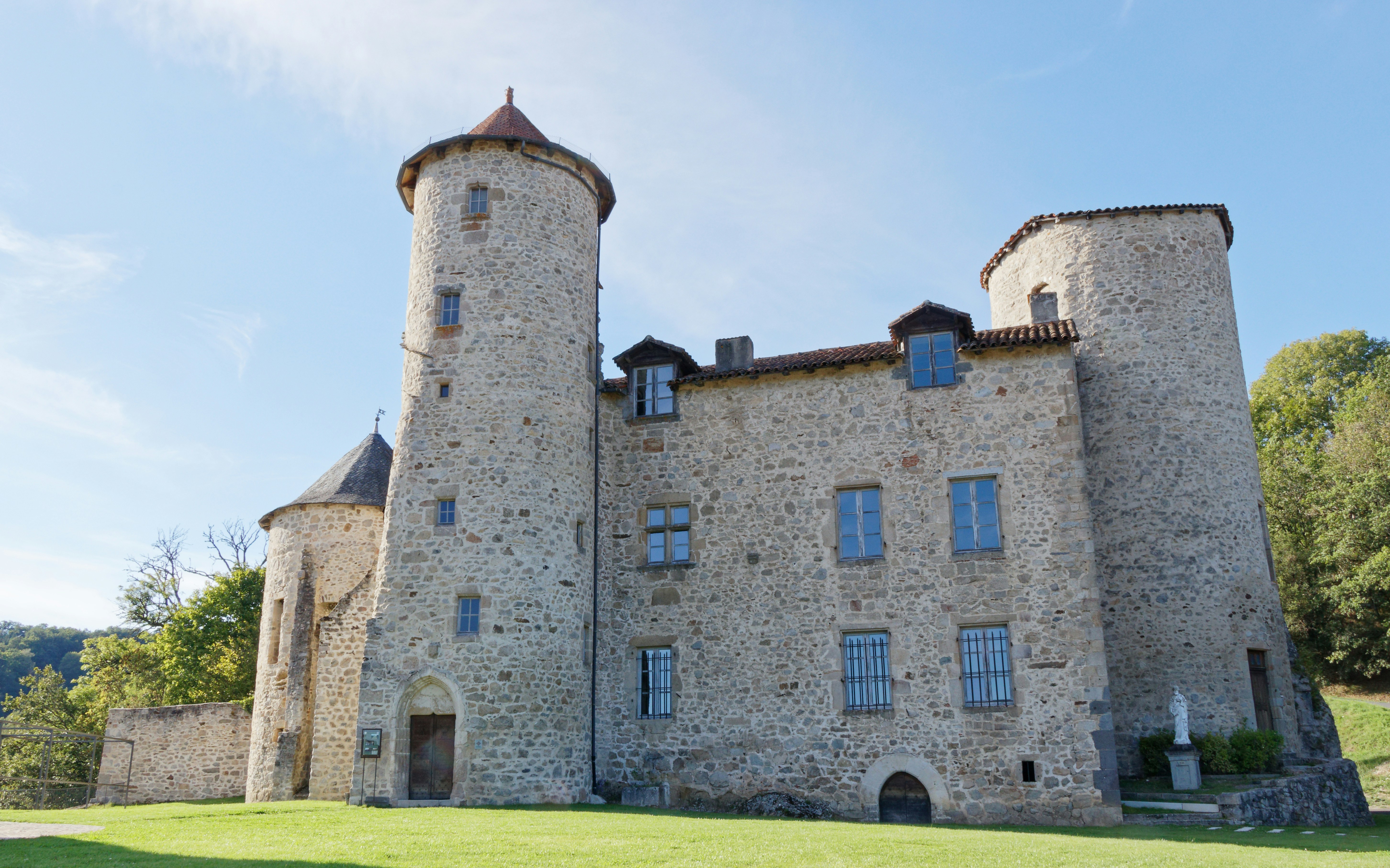 Historic stone castle featuring cylindrical towers and a lush green lawn, surrounded by trees under a clear blue sky.