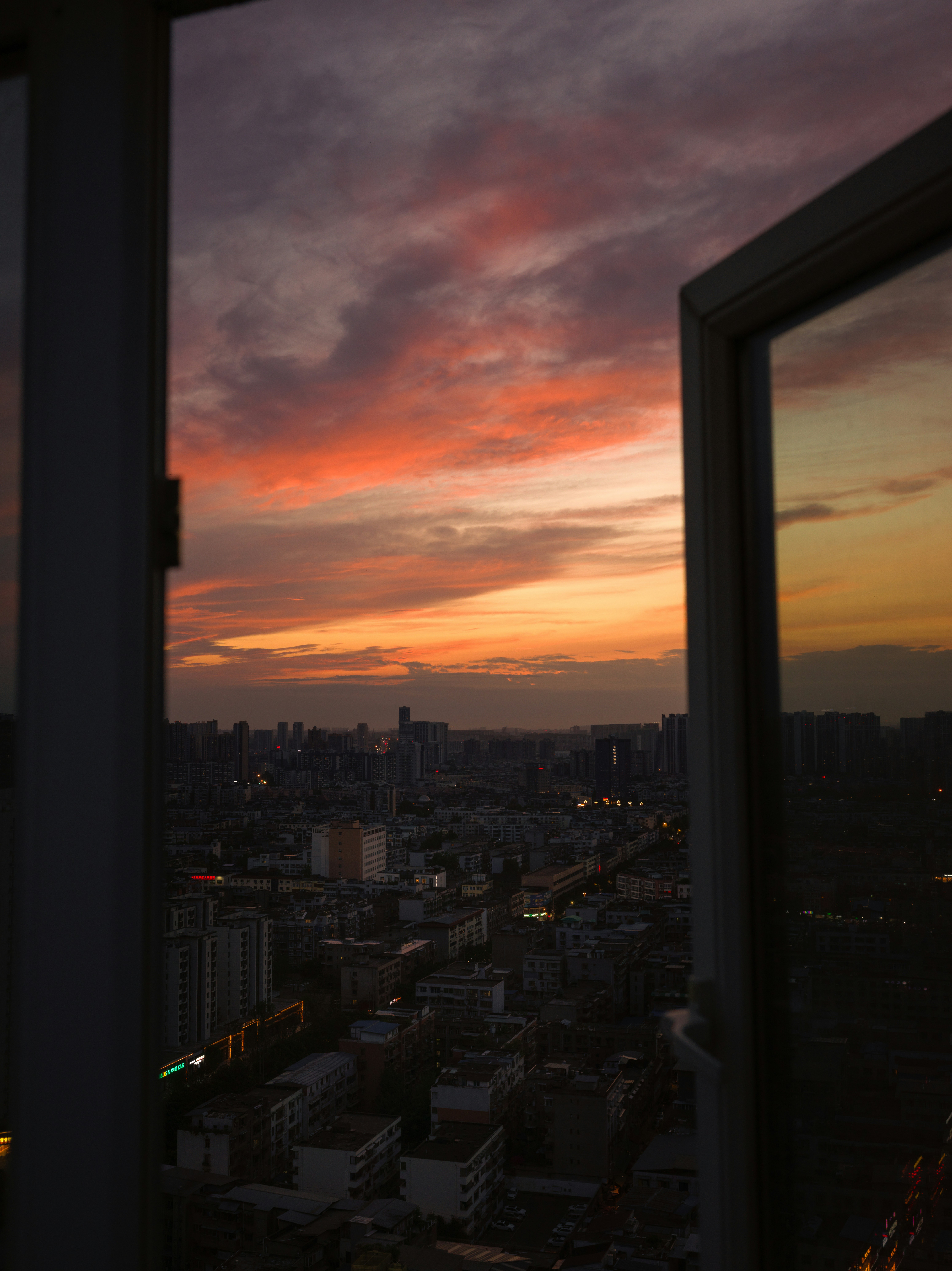 Vibrant sunset viewed through an open window, showcasing a cityscape bathed in warm hues. The scene captures the transition from day to night.