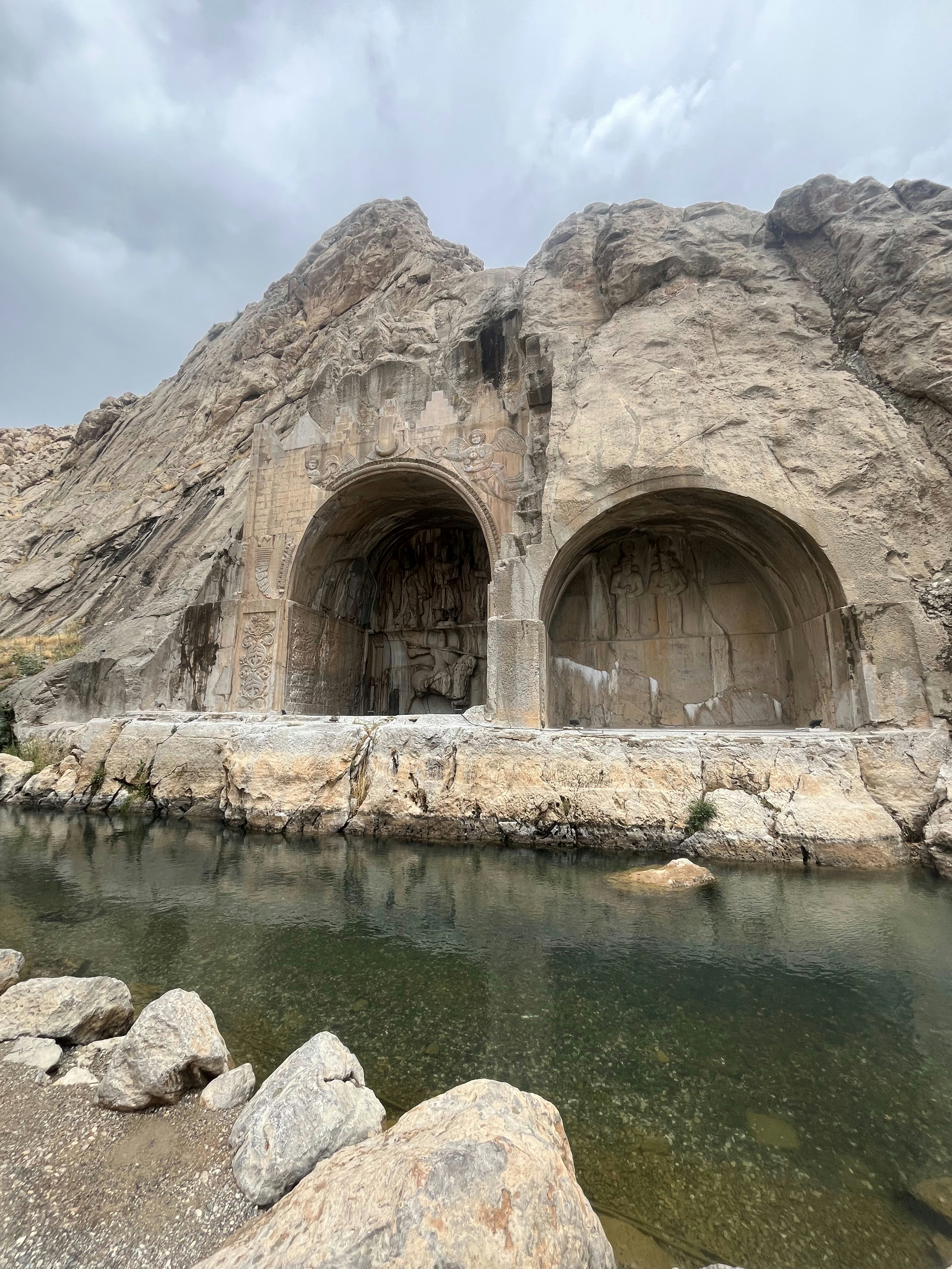 Intricate carvings on a rocky cliffside near a serene body of water, showcasing historical artistry and natural beauty.