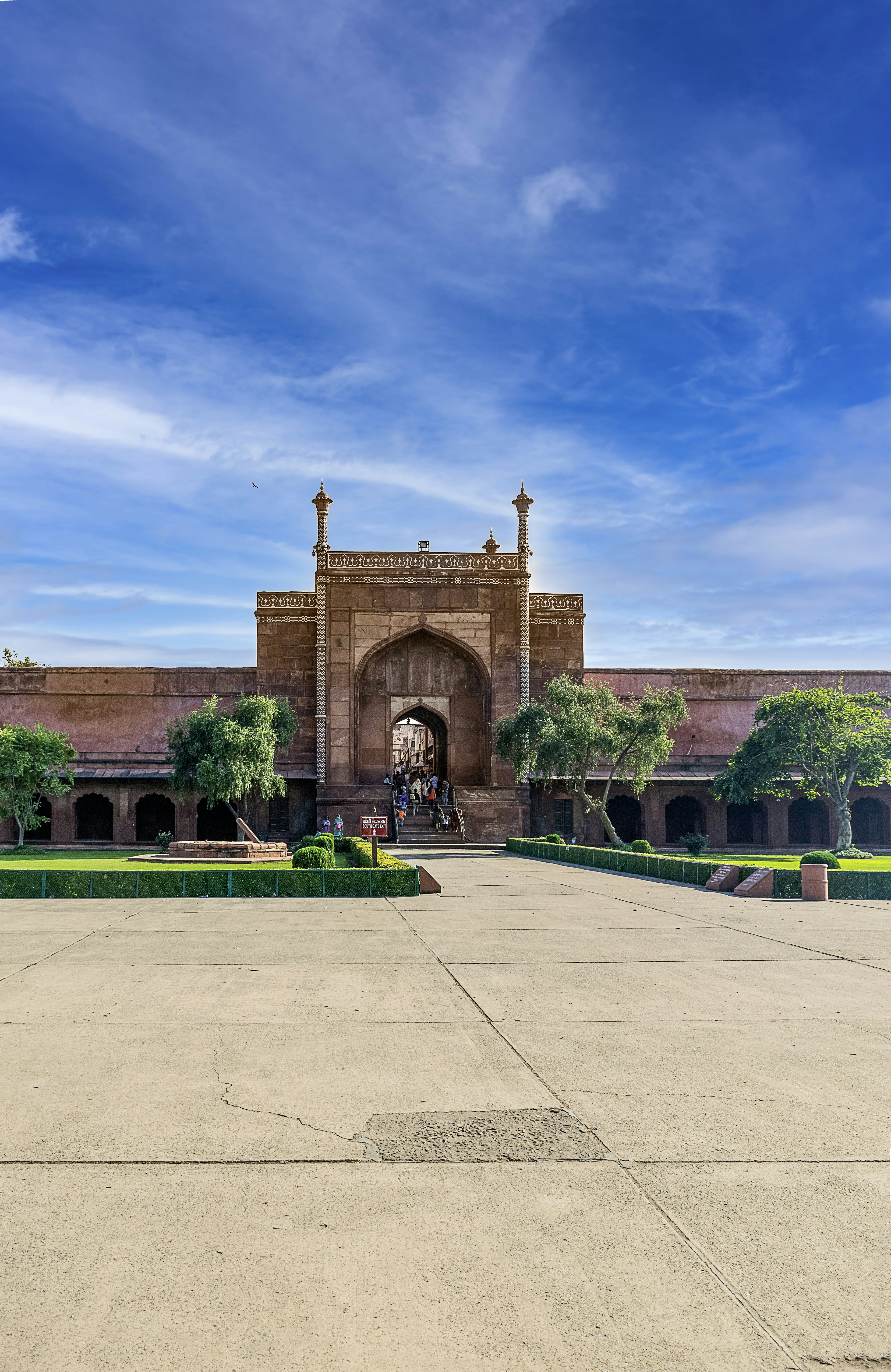 Majestic archway leading into a historical site, framed by lush greenery and a clear blue sky. Visitors gather at the entrance, hinting at the rich stories within.