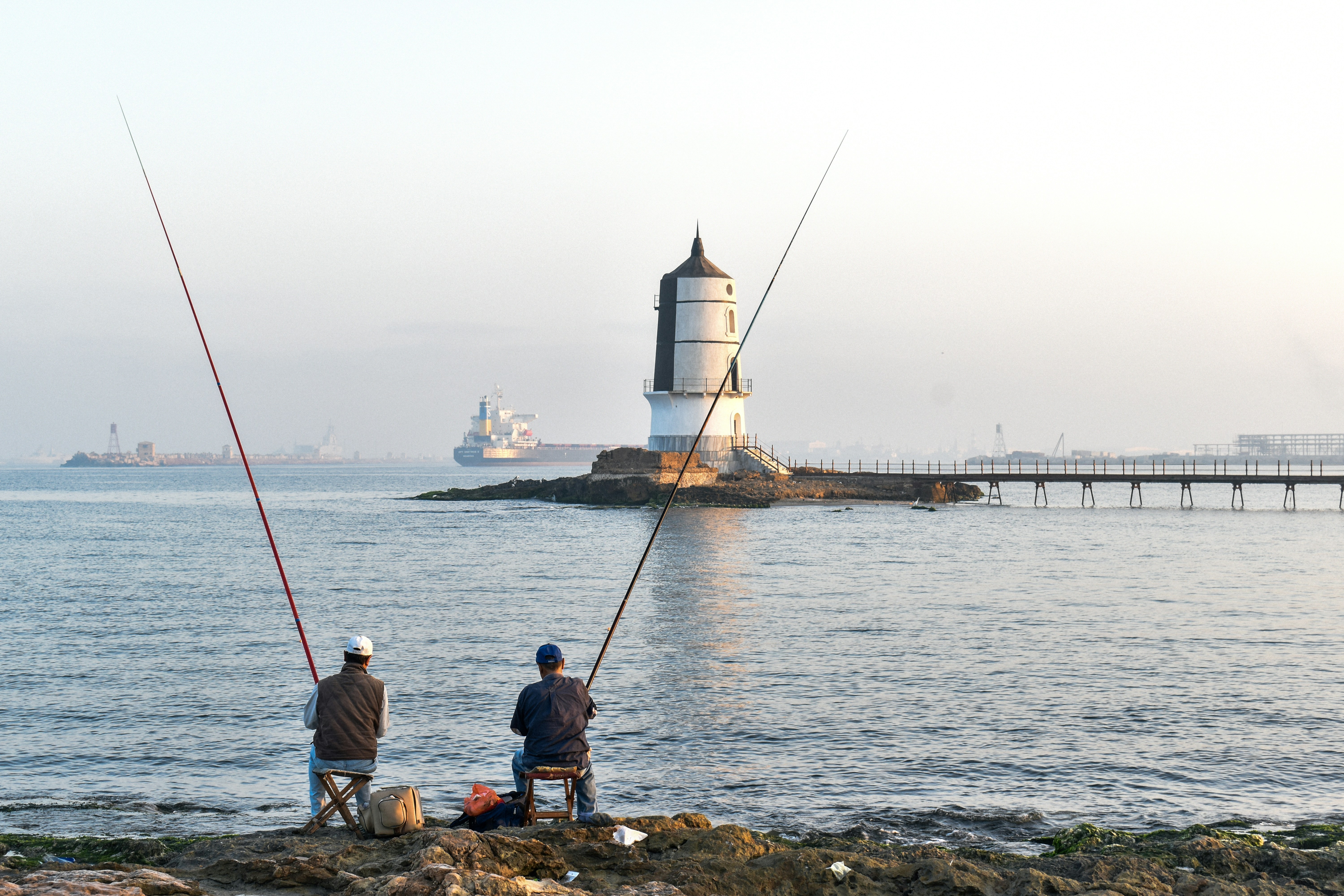 Two fishermen casting their lines near a coastal lighthouse at dawn, with a cargo ship visible in the distance.