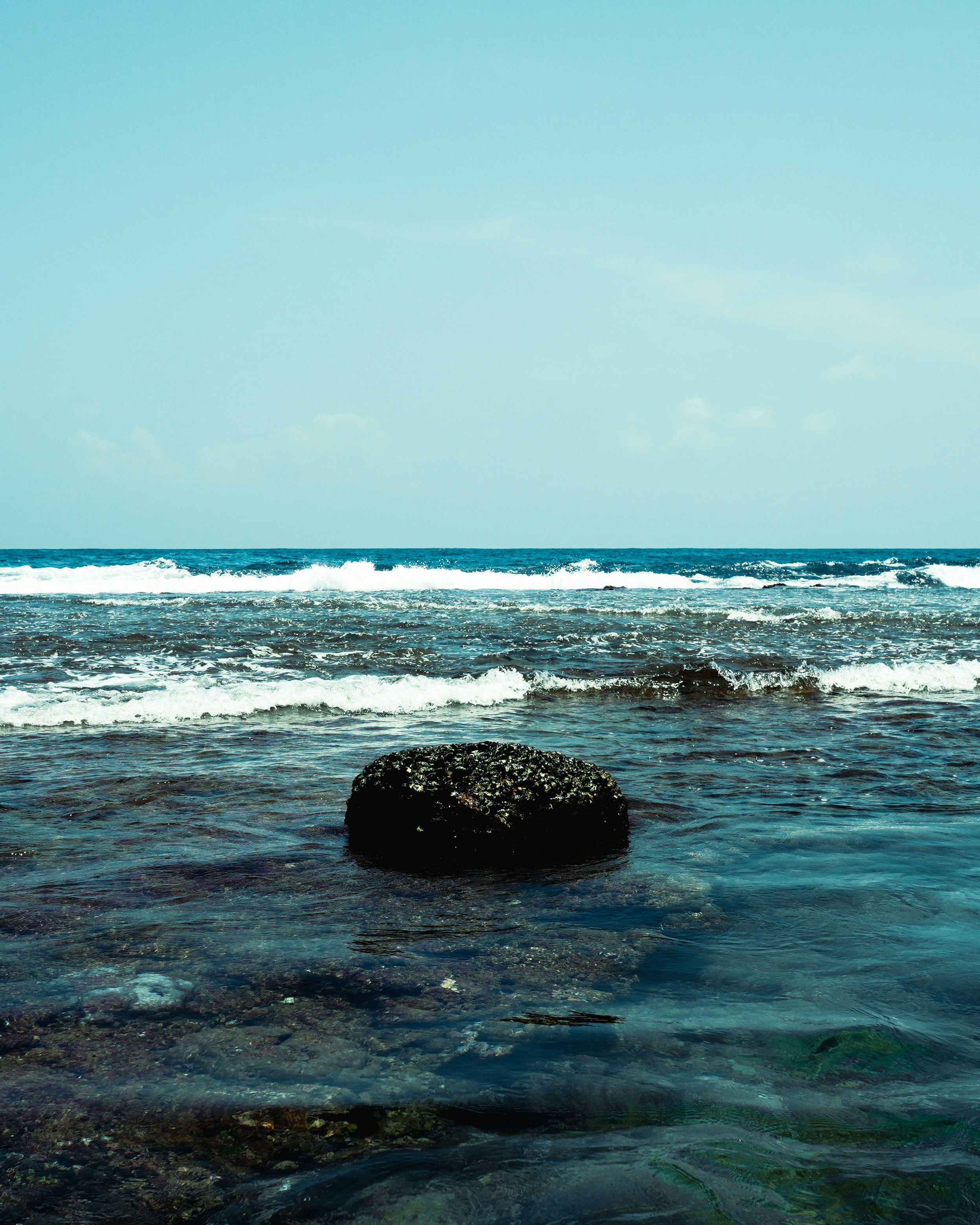 Lone rock emerging from clear waters, framed by gentle ocean waves under a bright sky.