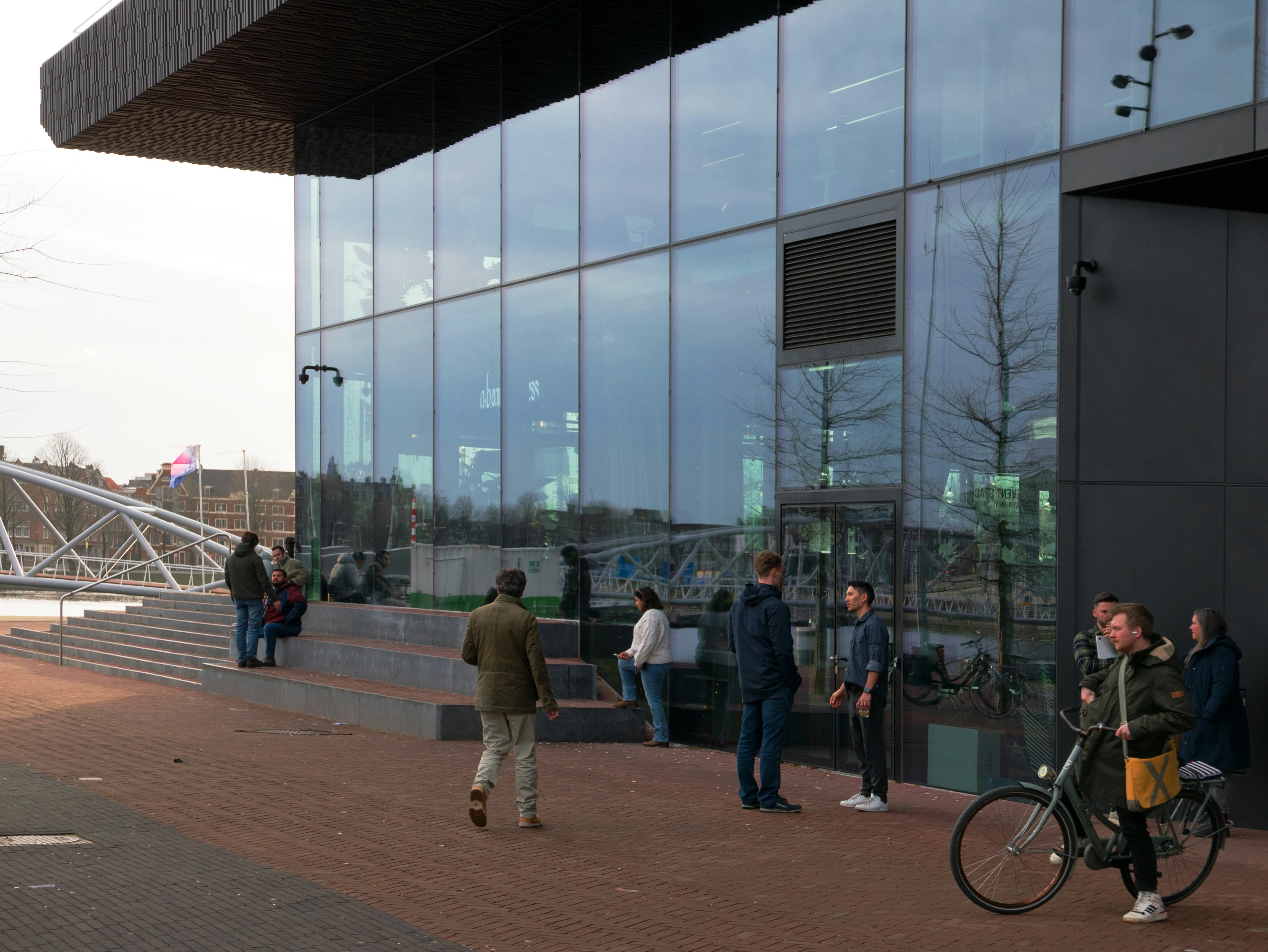 Free photo of people in front of the glass wall facade of the office of Booking.com, at the Oosterdokseiland in Amsterdam city. The glass wall has transparency so you can see partly the interior of the building with its pillar construction. Urban street photography of people and street life in the cities of The Netherlands; free images by Fons Heijnsbroek, 2023 city wallpaper in free download. / Foto van jonge mannen die voor de glazen wand van het kantoor van Booking.com op het Oosterdokseiland in Amsterdam staan en lopen. De moderne architectuur van het gebouw heeft veel glazen wanden.