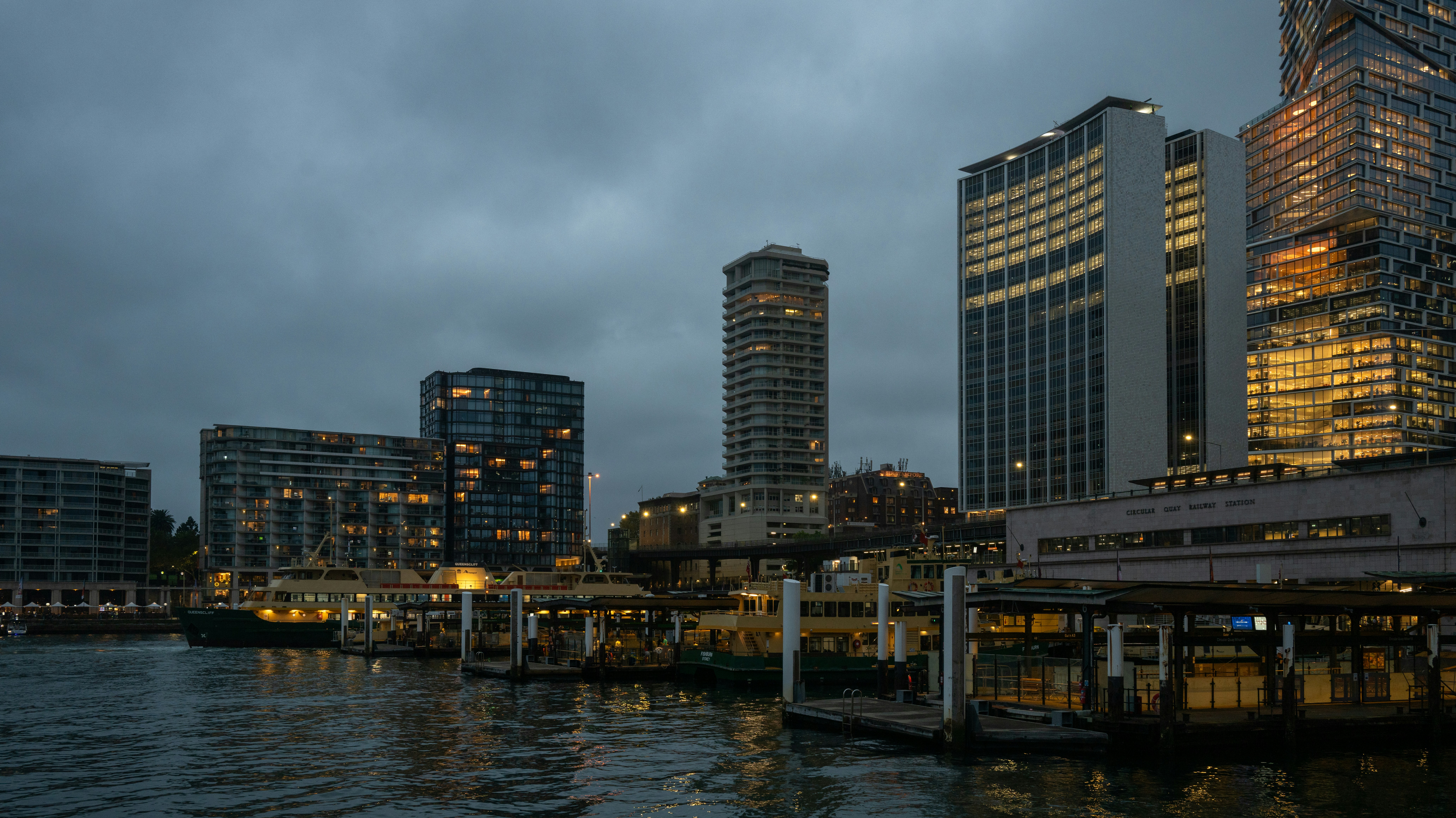 City skyline reflecting on water during twilight, with illuminated buildings and a ferry terminal in the foreground.