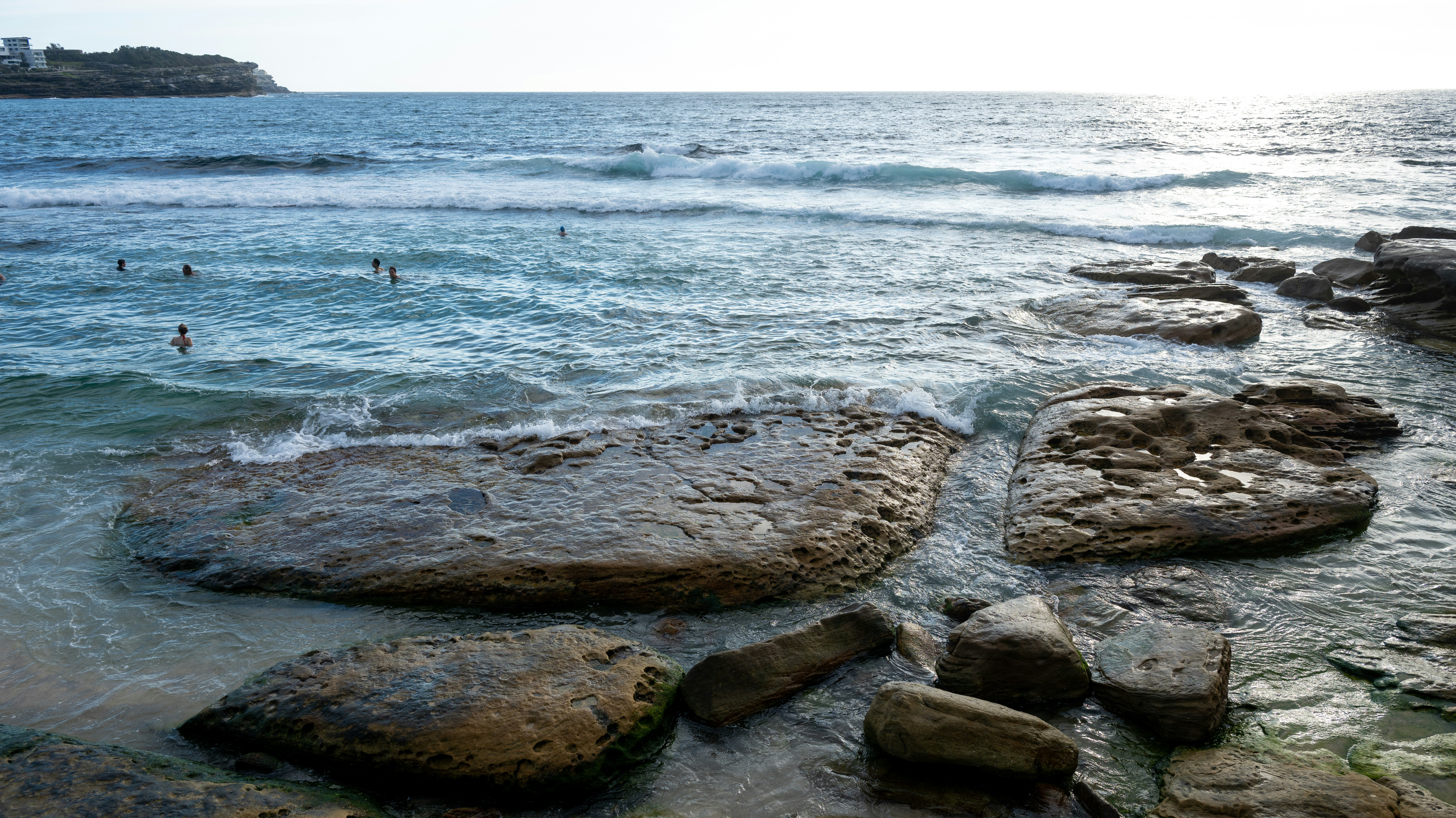 Waves gently lap against textured rocks at a tranquil beach, with swimmers enjoying the cool waters. The horizon meets the sky in a serene coastal scene.