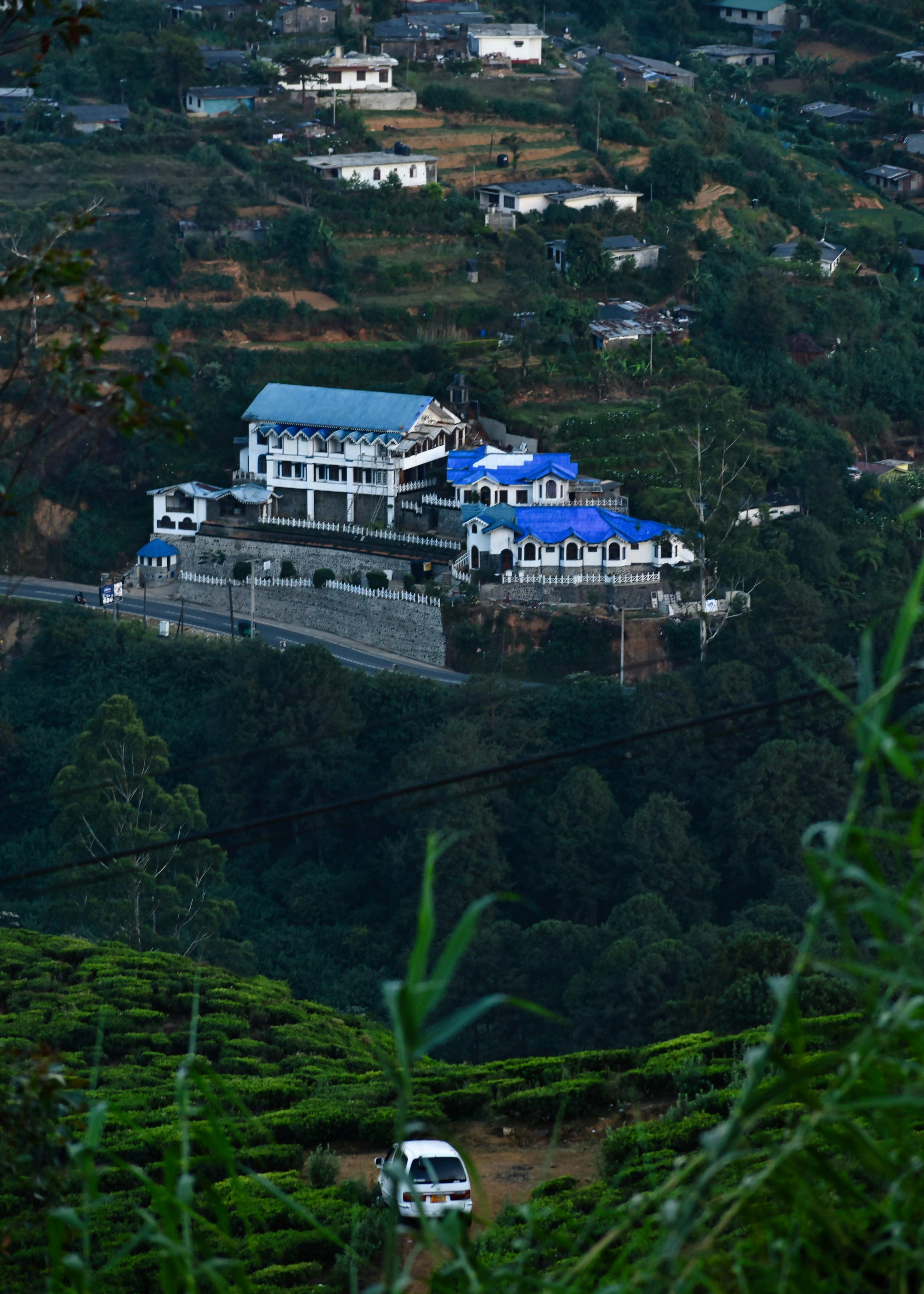 A picturesque hillside retreat featuring a blue-roofed building surrounded by lush greenery and tea plantations. The winding road adds to the tranquil atmosphere.