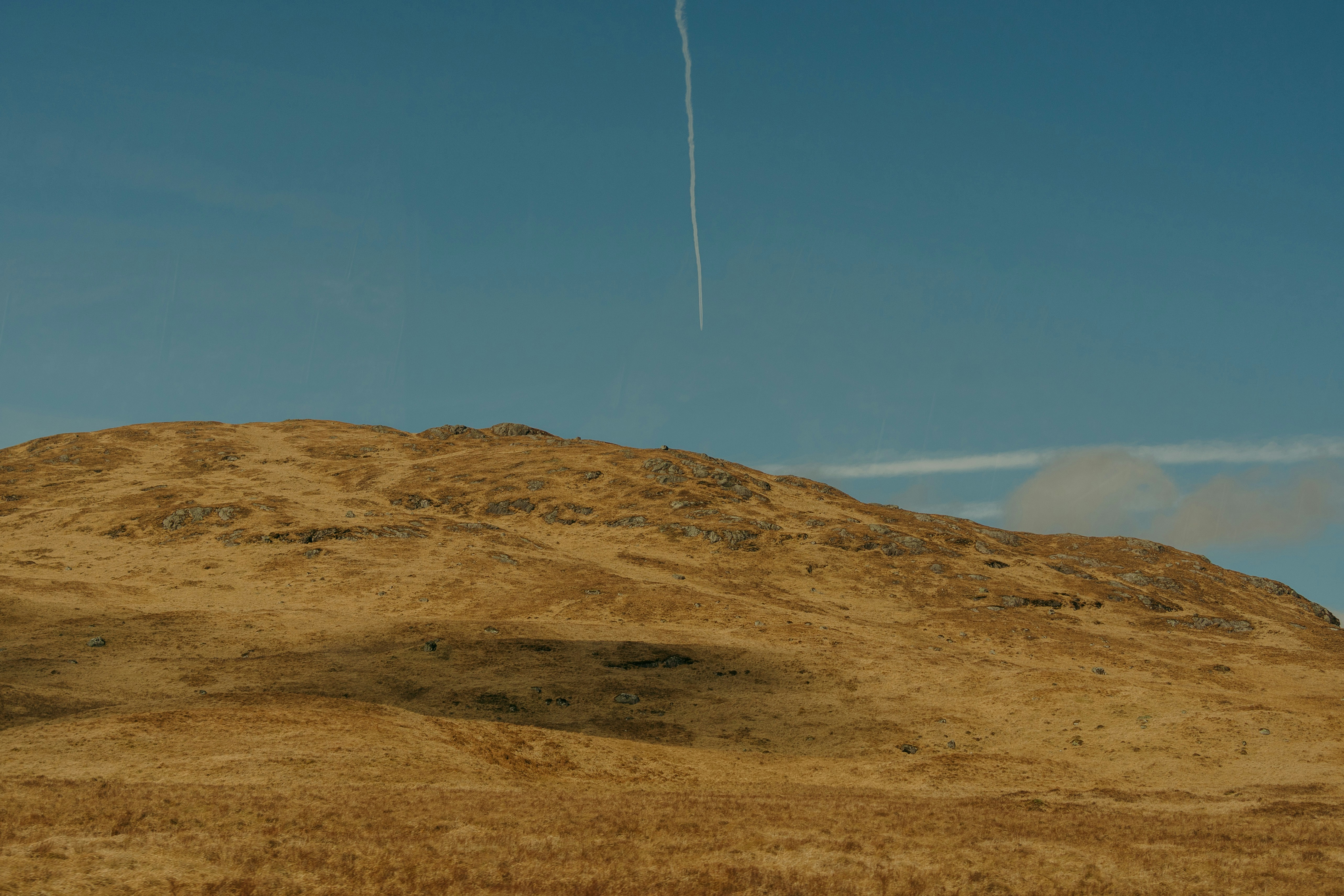 Golden hills stretch under a clear blue sky, marked by a faint contrail cutting through the tranquility. A subtle interplay of light and shadow enhances the natural beauty.