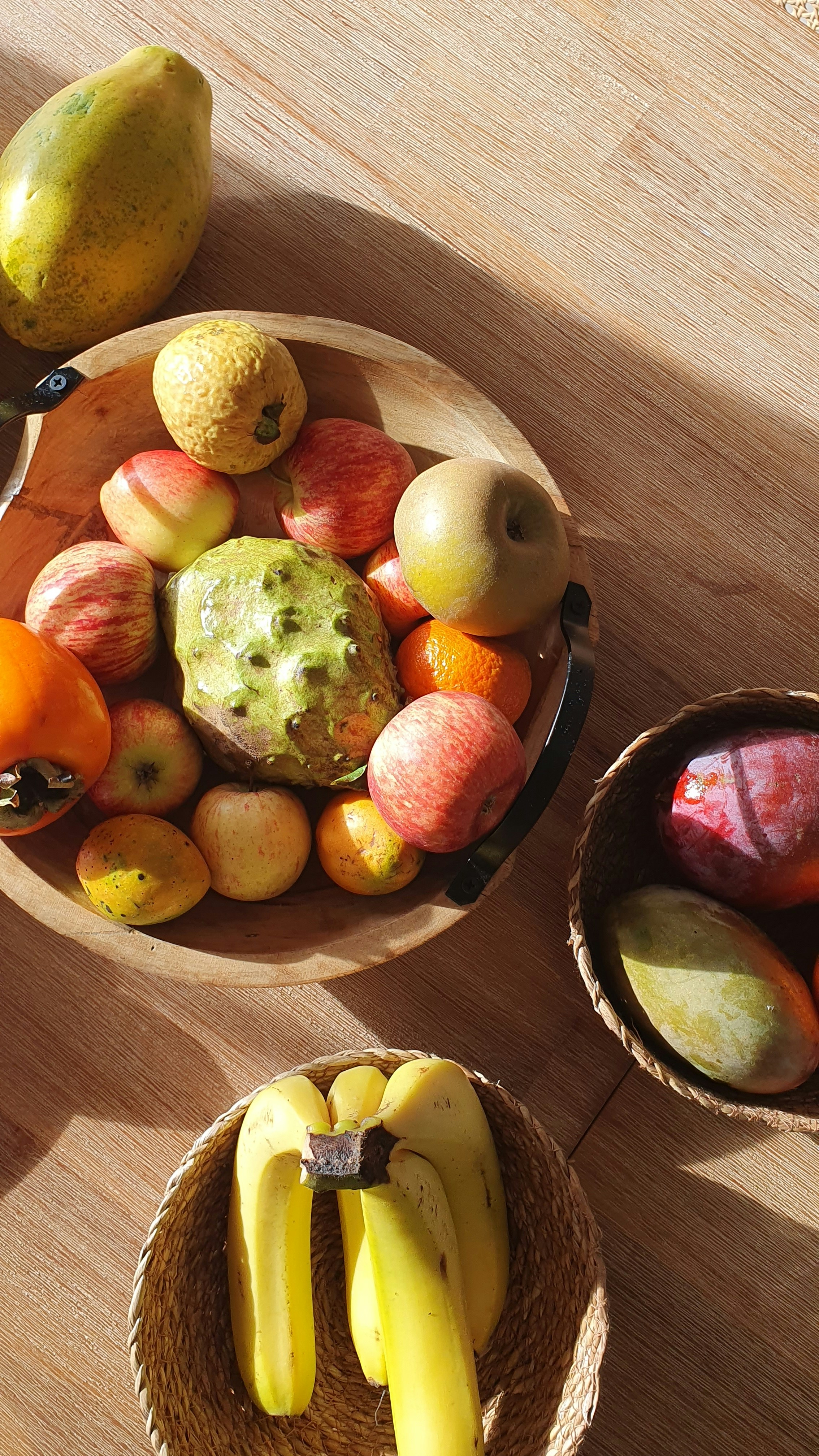 Sunlit still-life photograph of a wooden table with fruit bowls, centered on a spiky jackfruit surrounded by apples, pears, and bananas.
