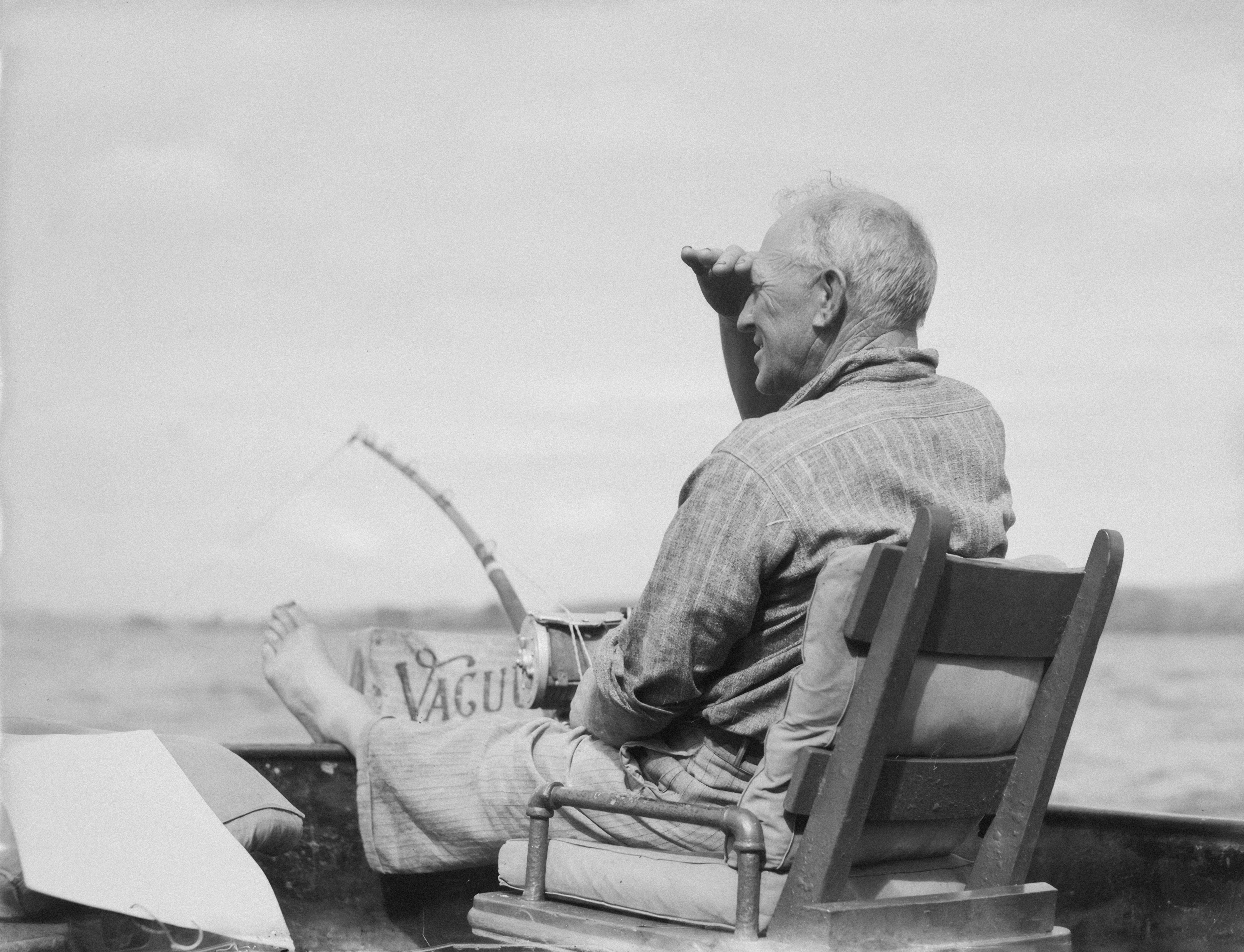www.aucklandmuseum.com/discover/collections/record/1054972 [Man fishing from a chair on a boat] This photograph was taken by Tudor Washington Collins (b.1898, d.1970), possibly in the 1940s. This back-side view shows a man seated on a padded chair on a fishing boat, holding a fishing rod, holding a hand to his forehead and appearing to be overlooking the sea, positioned casually with his left leg raised onto the boat's edge, where a piece of wood with the letters "VAGU" visible is placed. Credit: Shared by Auckland War Memorial Museum, Tāmaki Paenga Hira, as part of the Tudor Collins collection. Rights: No known copyright restrictions. Reference: