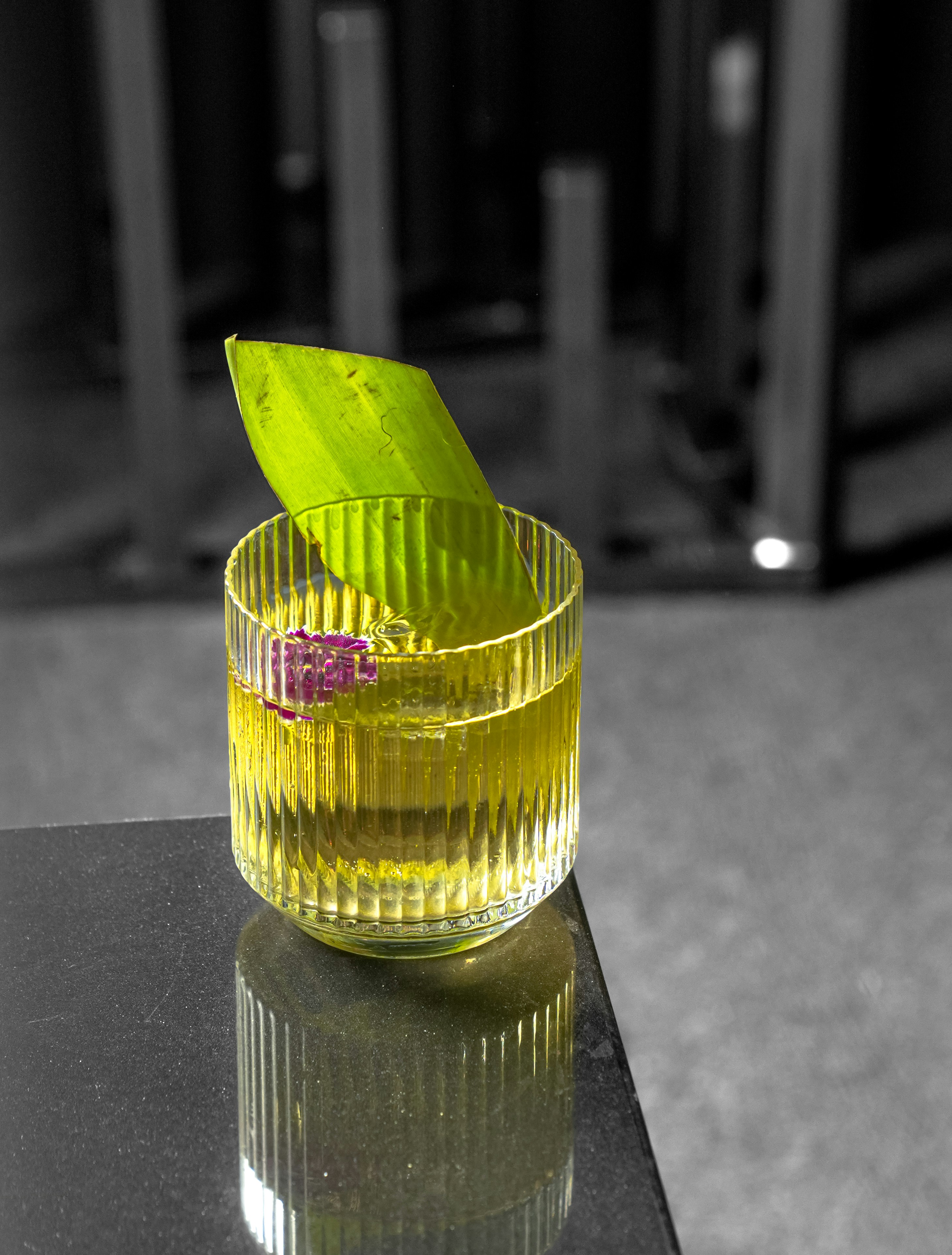 Vibrant cocktail garnished with a fresh aloe leaf and a delicate flower, resting on a sleek black surface. The background features a blurred bar setting.