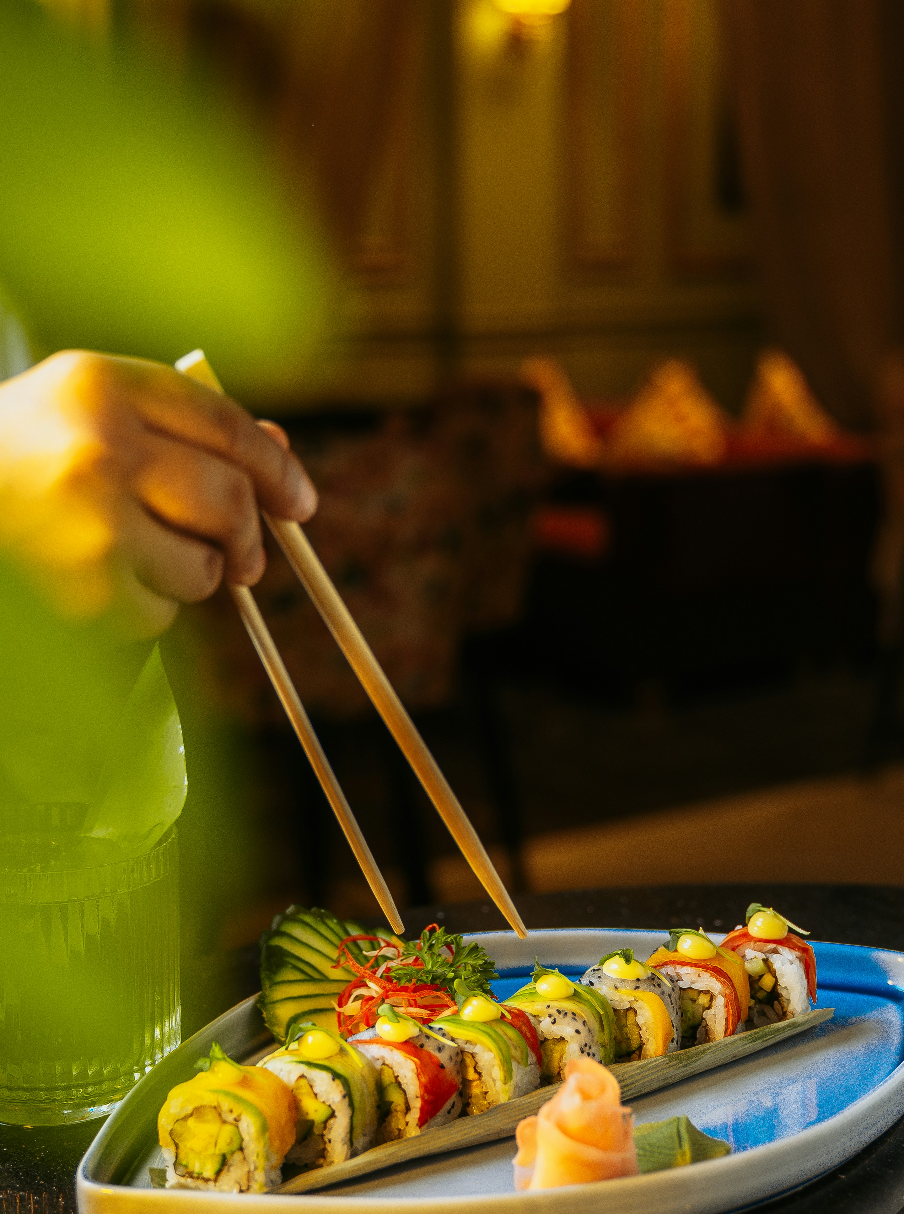 Colorful sushi rolls arranged on a plate with chopsticks poised for selection. A refreshing drink and a blurred background enhance the dining atmosphere.