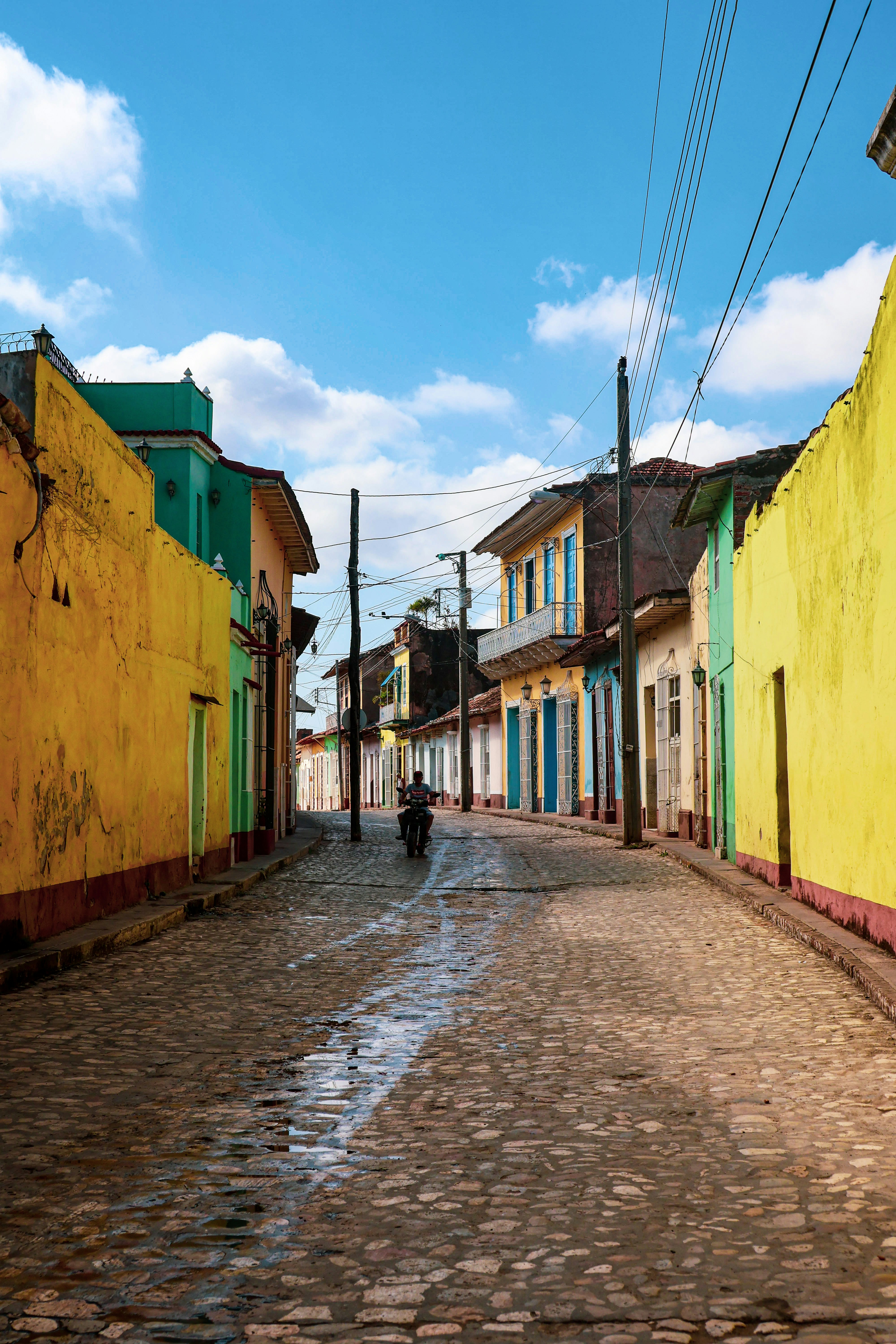Colorful colonial buildings lining a cobblestone street reflect the charm of a historic town. A lone cyclist navigates the winding path under a bright blue sky.