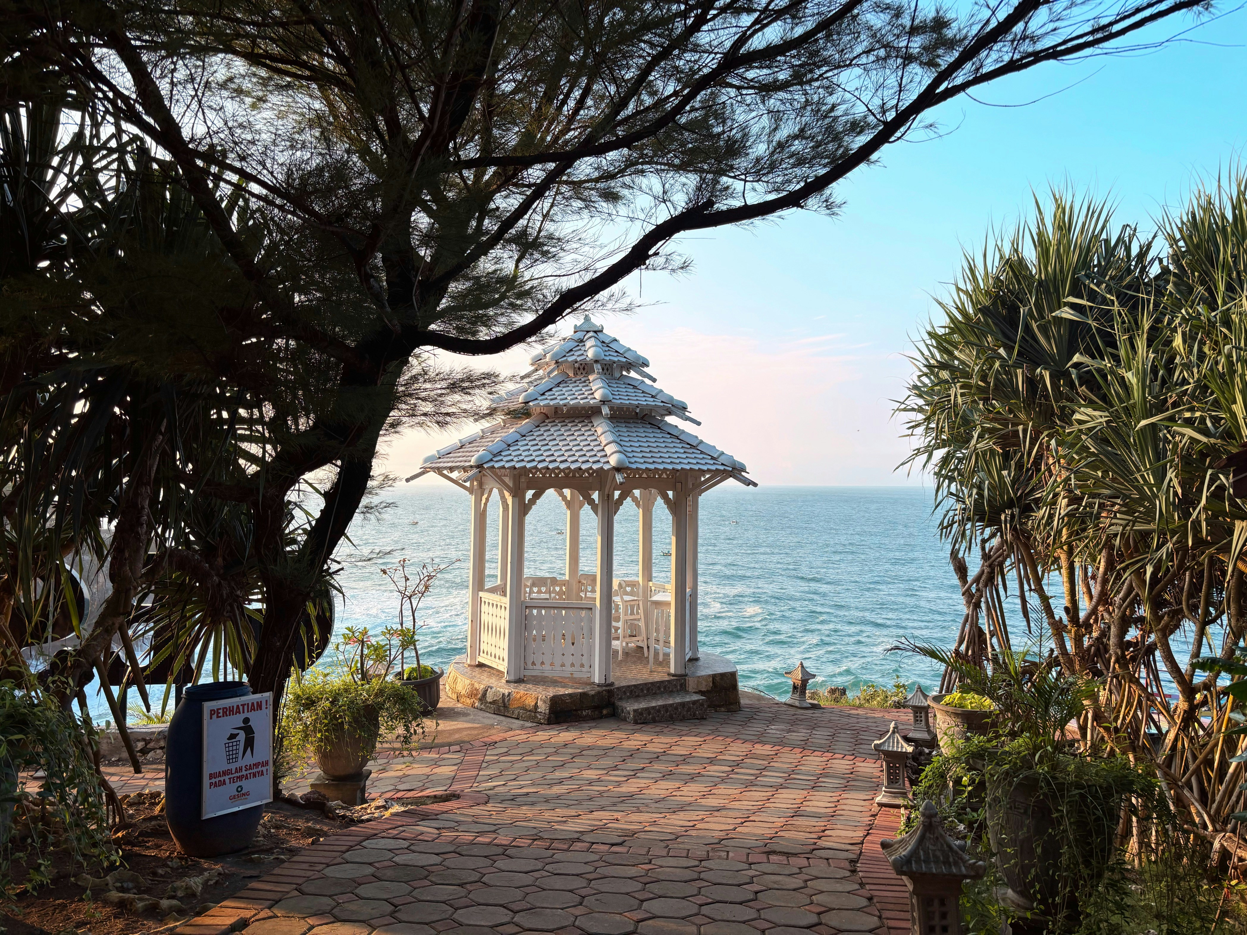 White seaside gazebo on a brick promenade framed by pines overlooking the sea