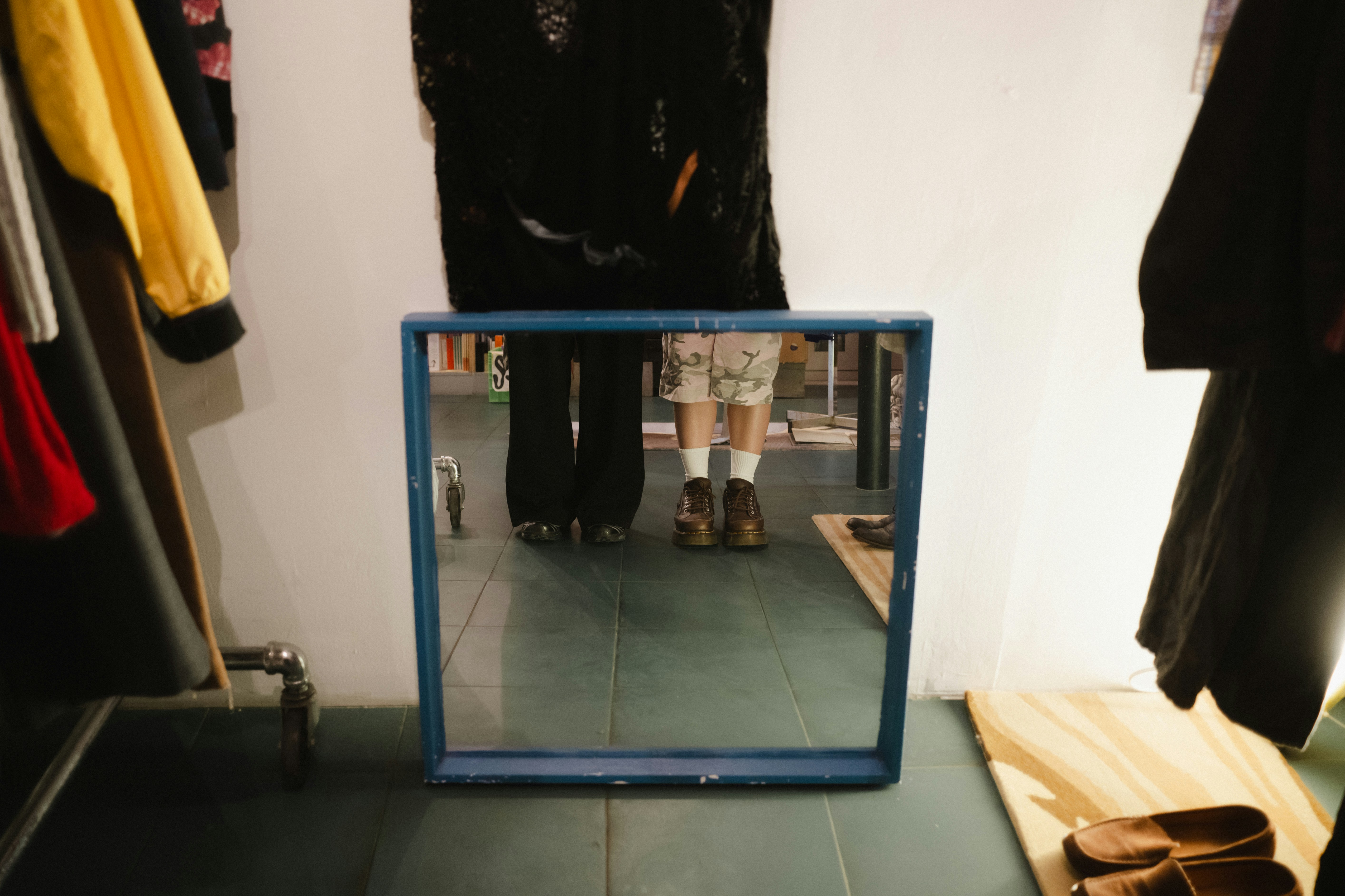 A mirror reflects the lower halves of two individuals standing amidst clothing, highlighting a blend of fashion and personal style.