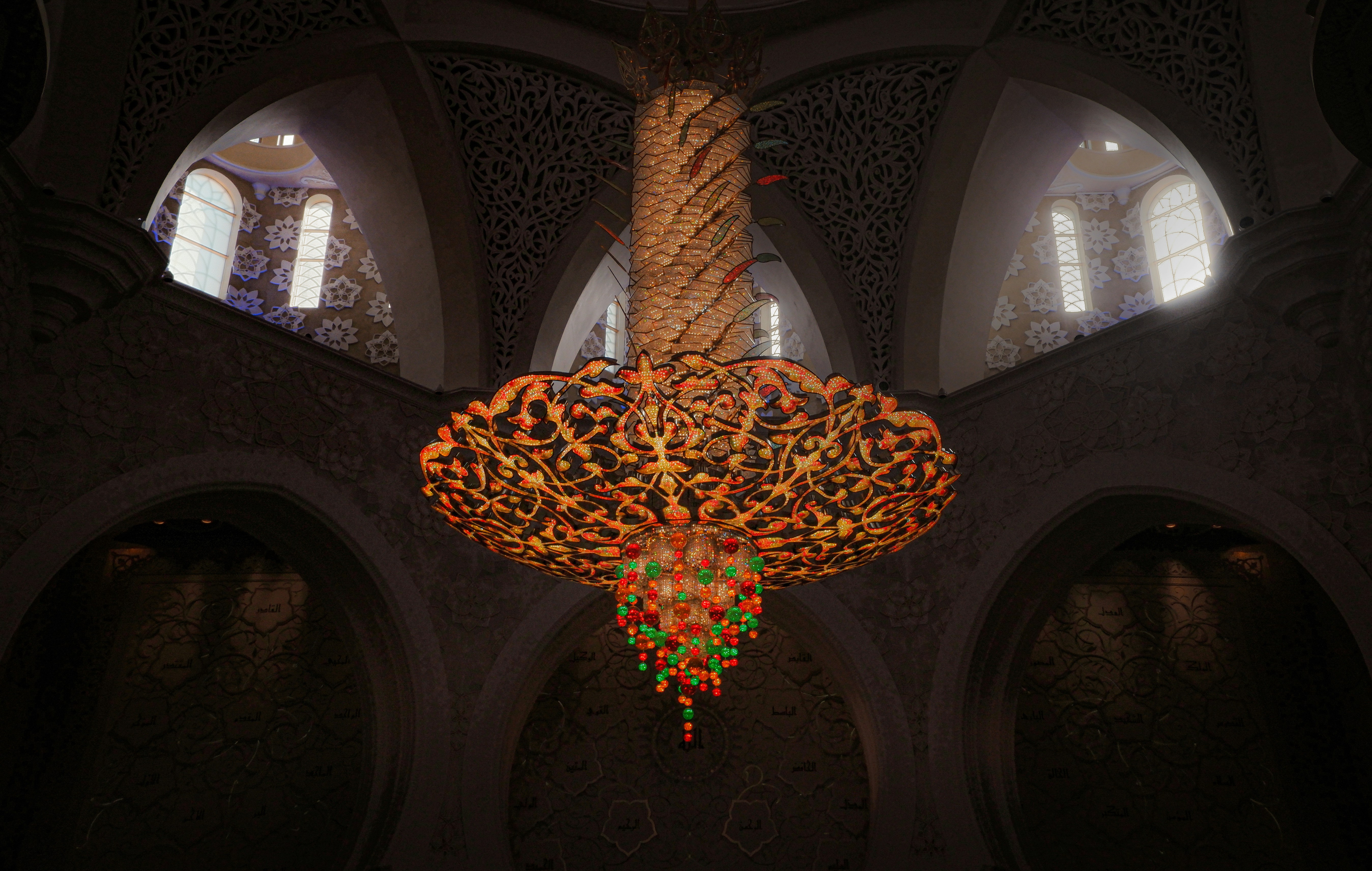 Intricately designed chandelier adorned with colorful crystals, suspended in an ornate architectural space with arched windows.