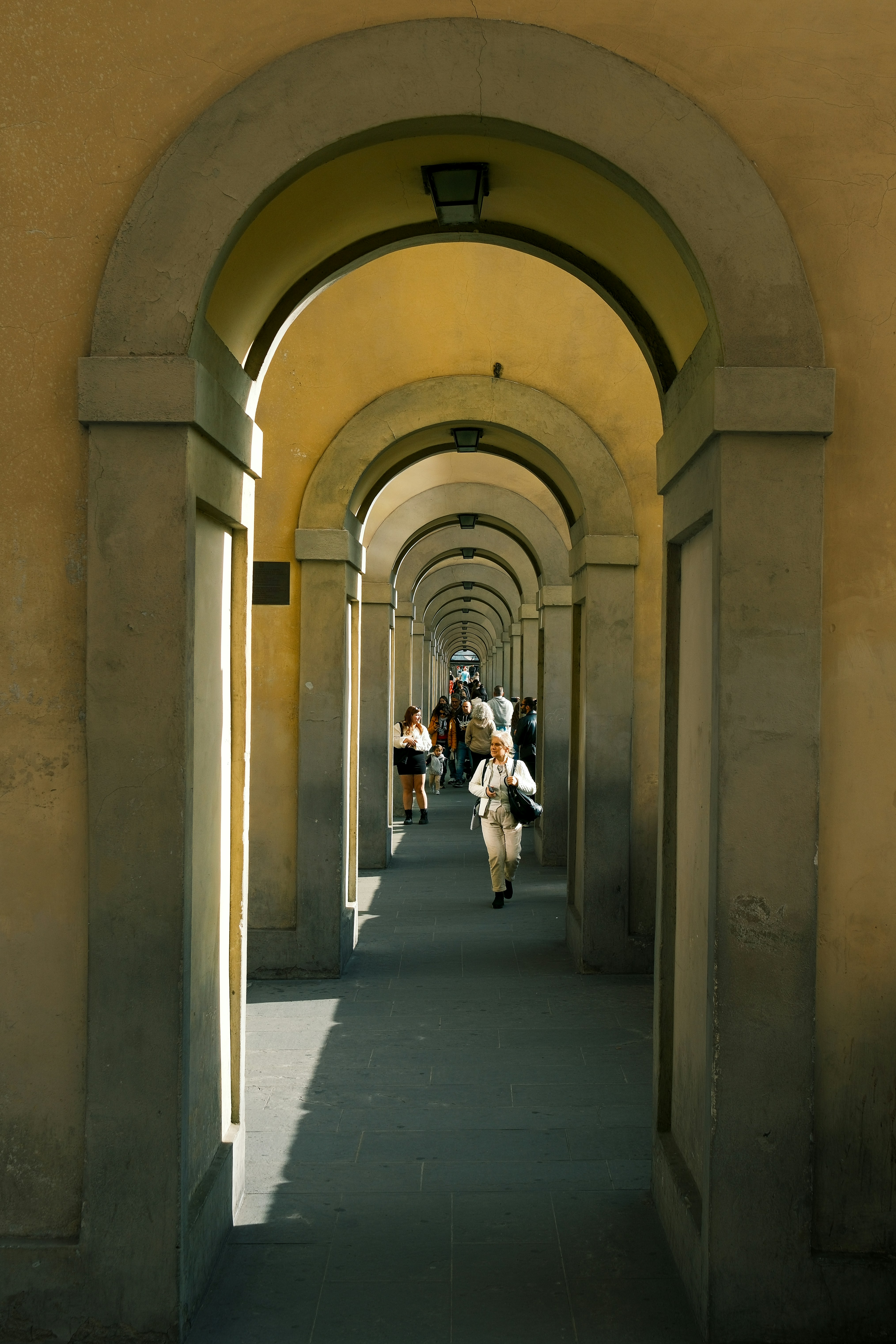 A line of arches in Florence leading to a crowd in the distance. Architectural symmetry and human flow. | People stroll down a long arched walkway.