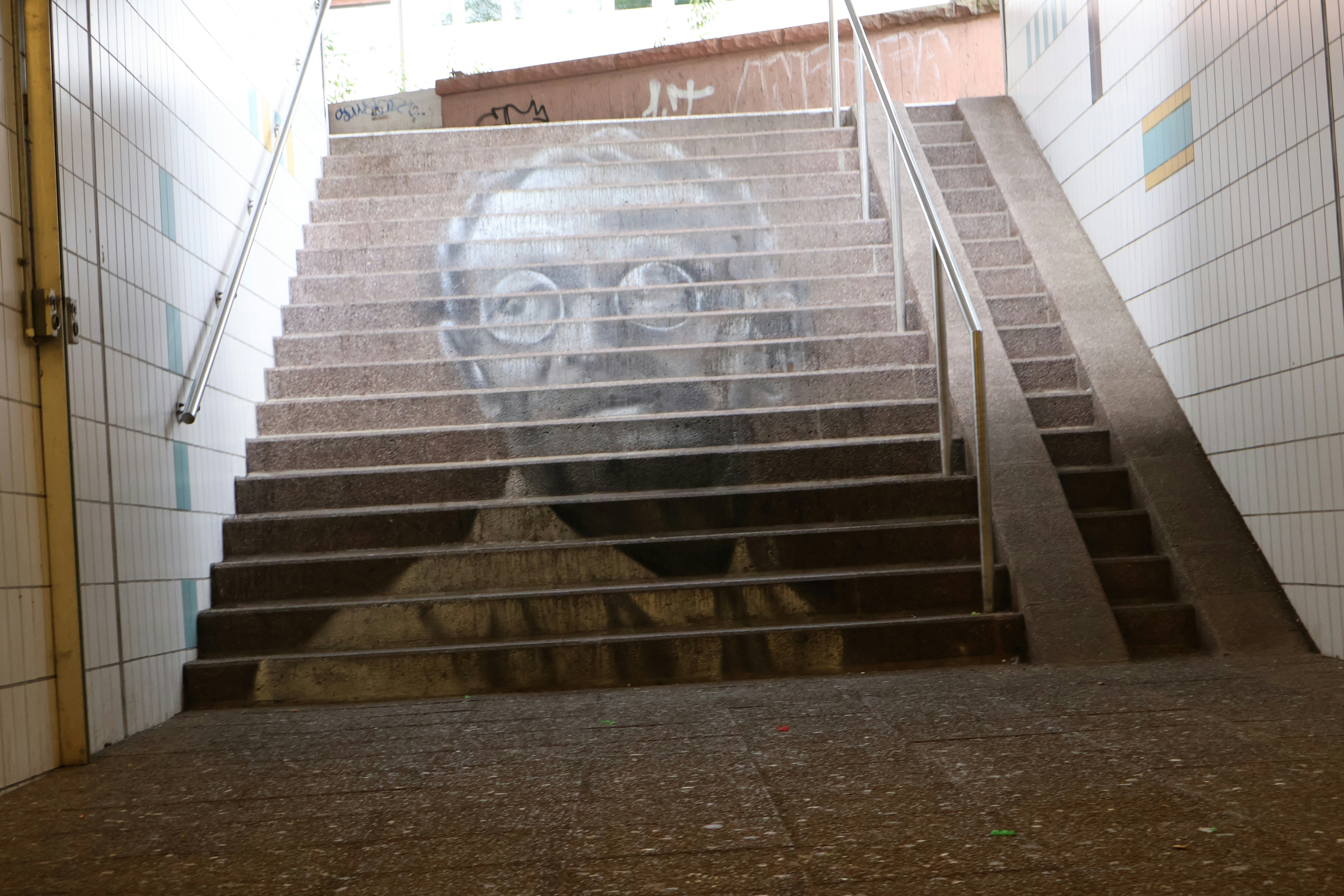 A large mural of a bespectacled man greets visitors on a staircase, blending art with urban architecture. The piece captures a moment in time, inviting reflection.