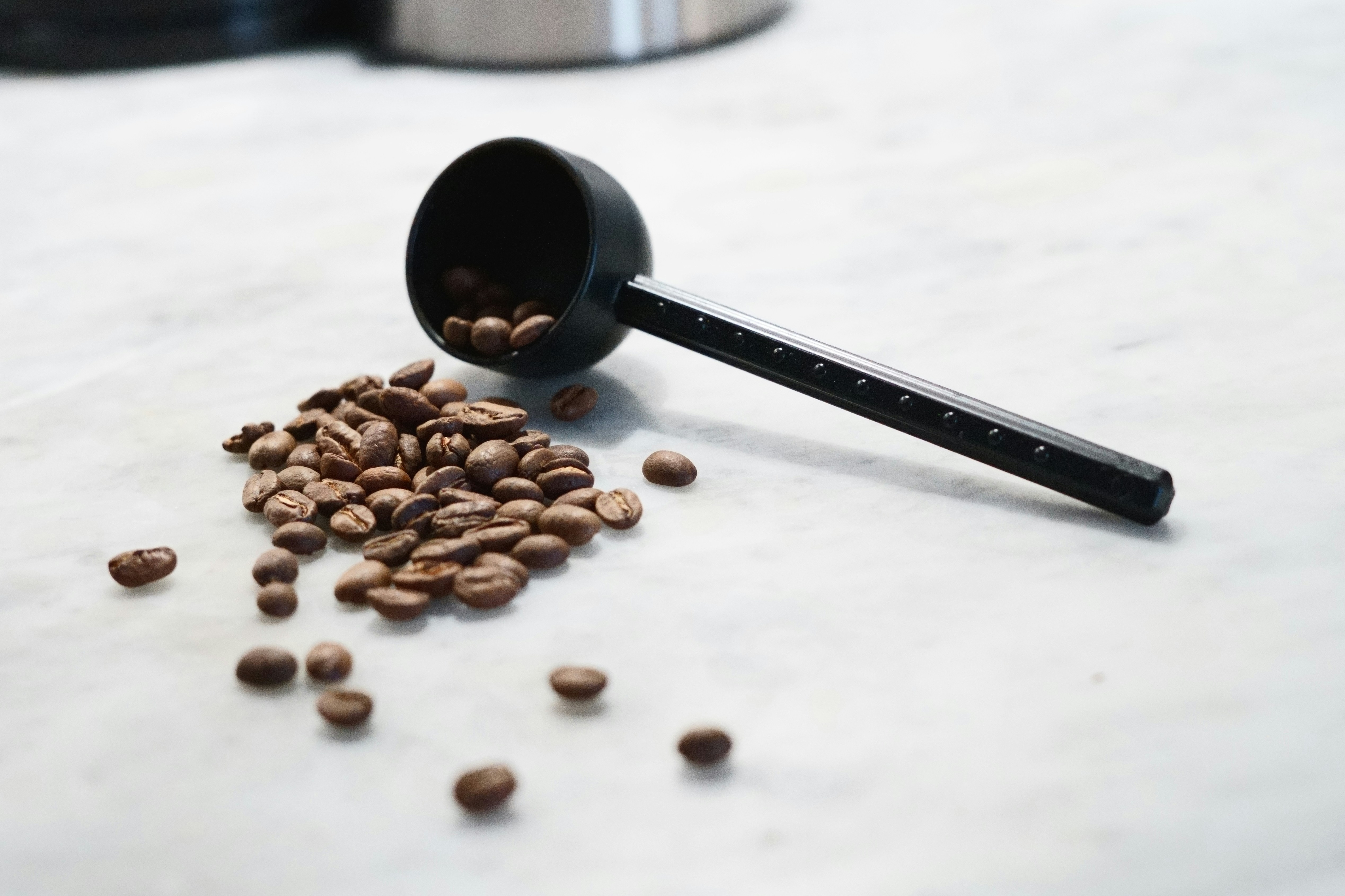 Black measuring scoop tipped over with roasted coffee beans scattered on a light marble surface.