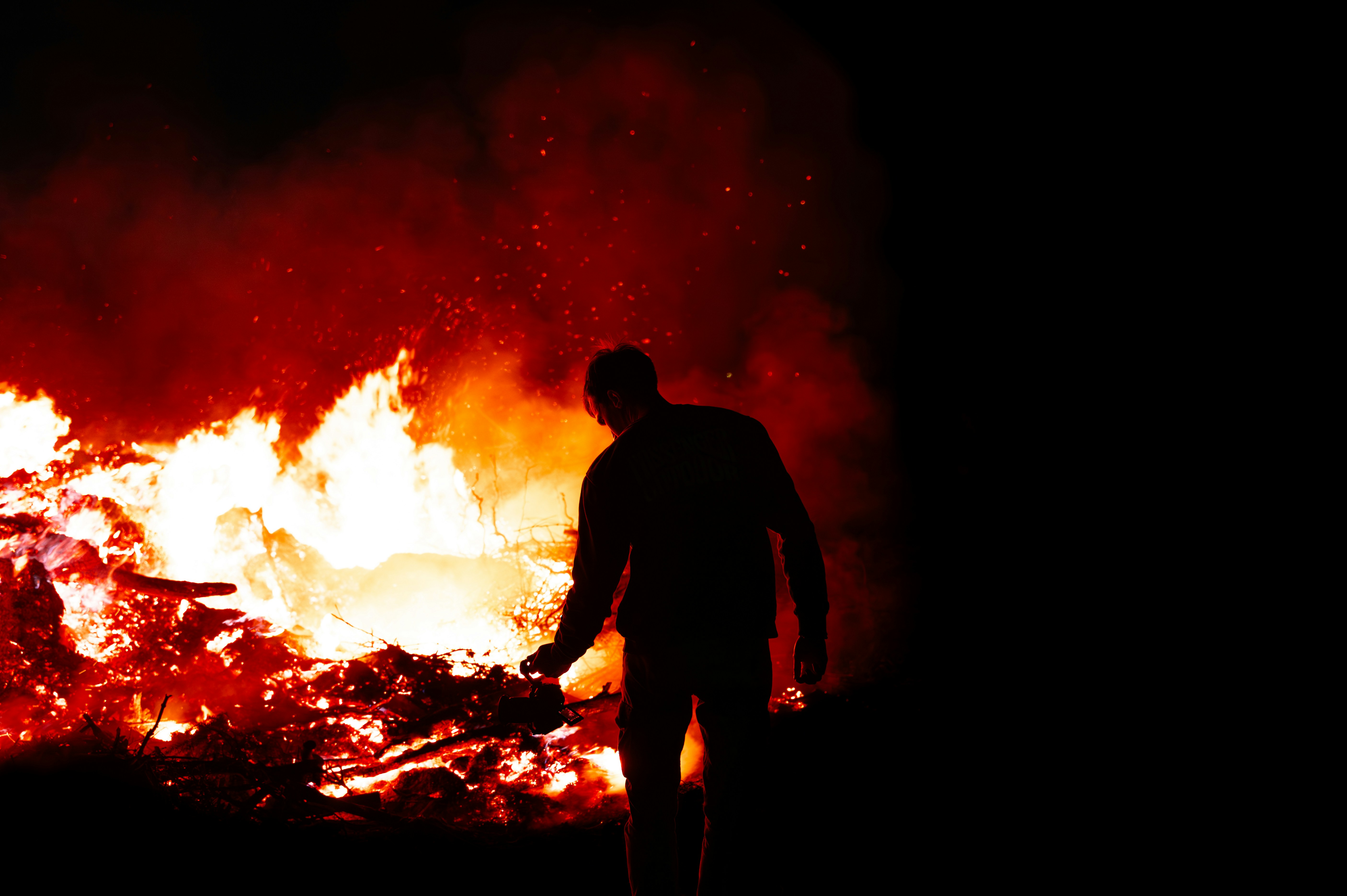 Silhouette of a person standing near a roaring fire, surrounded by glowing embers and smoke. The scene captures the intensity and raw energy of the flames.
