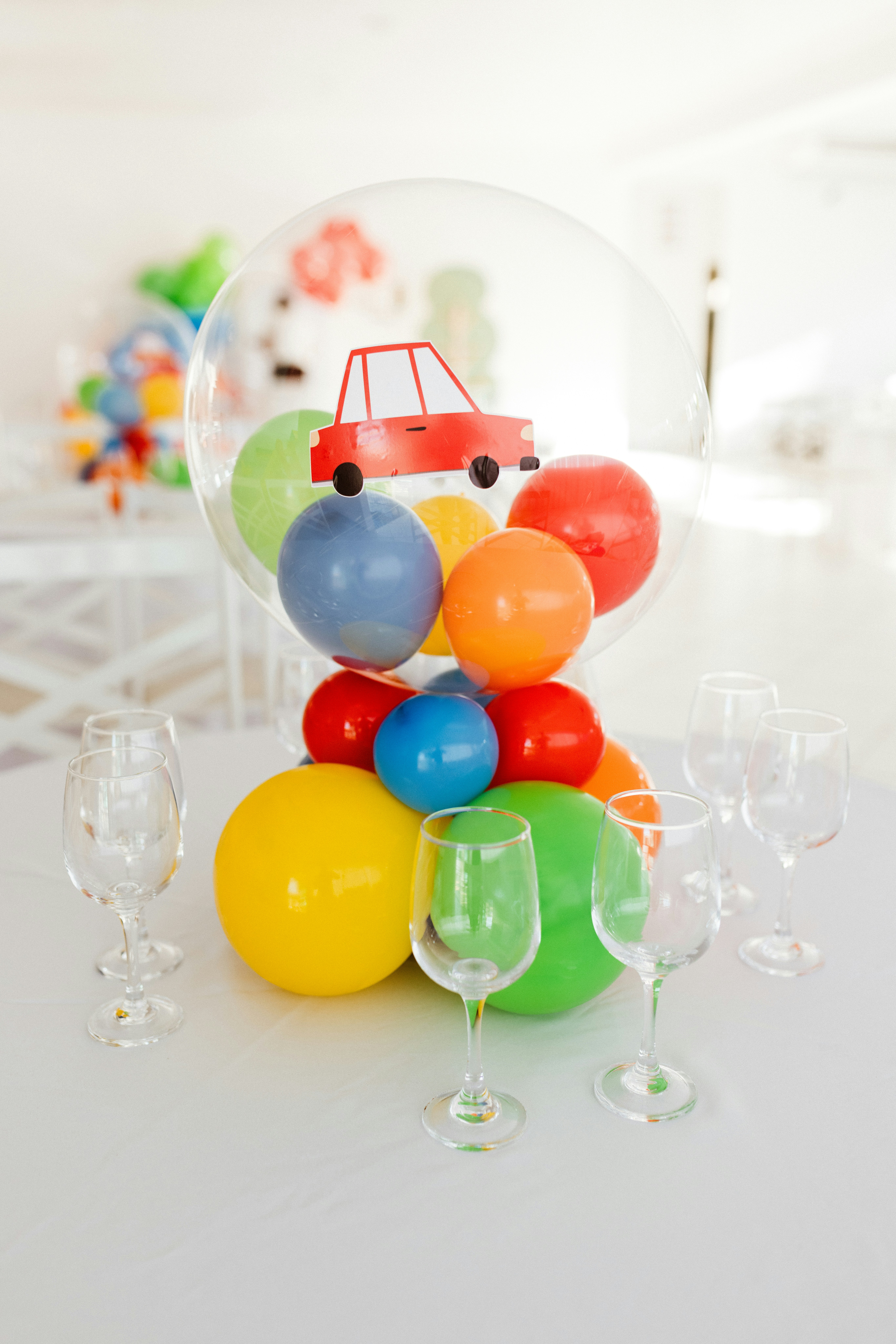 Colorful balloons stacked beneath a transparent dome featuring a cheerful red car. Elegant glassware surrounds the vibrant centerpiece.