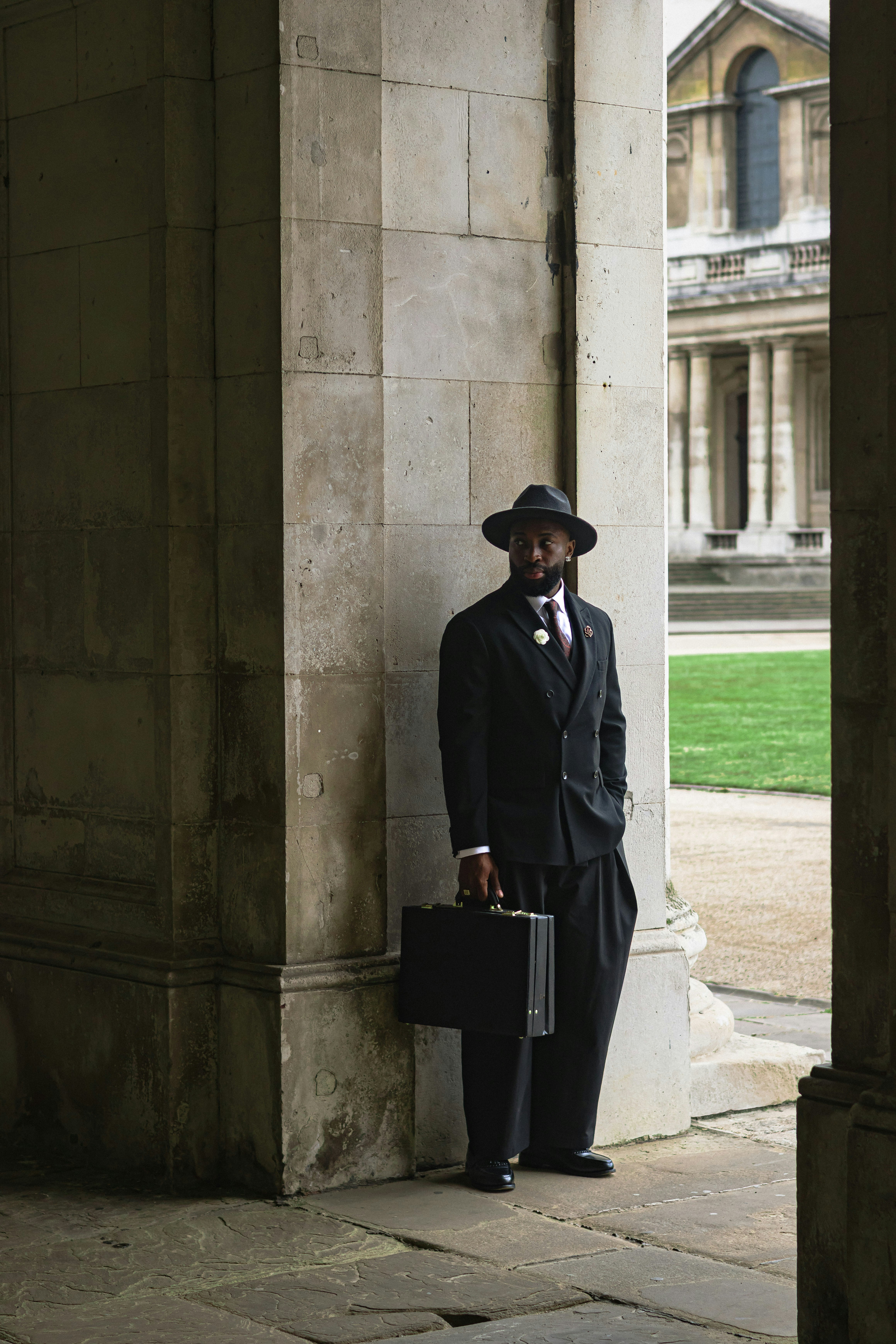A well-dressed man stands confidently in a stone archway, holding a briefcase, with a historic building in the background.