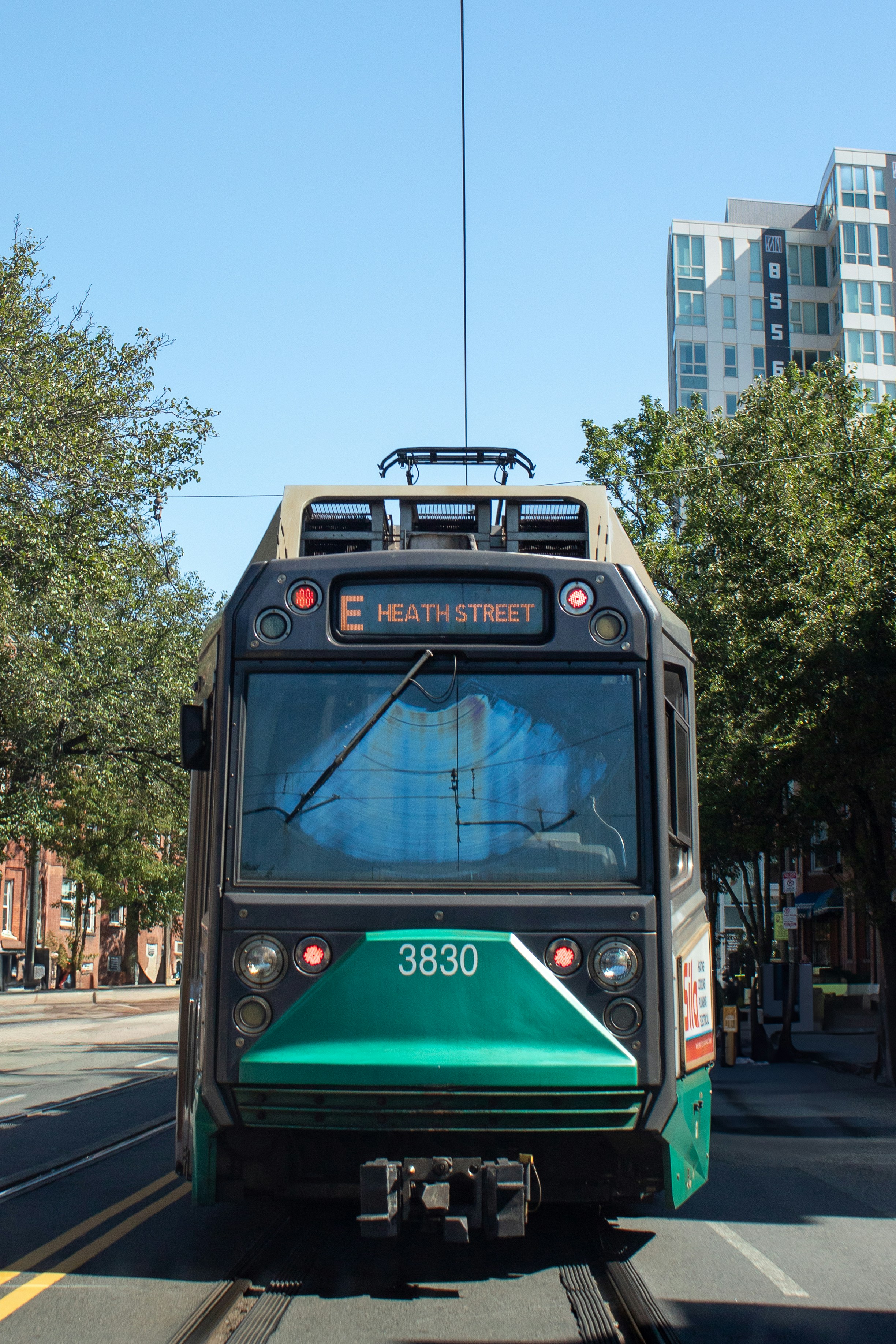 A front-facing view of a Boston Green Line E branch light rail train bound for Heath Street. The scene captures the blend of public transportation and urban greenery, set against a backdrop of modern apartments and a clear blue sky. Perfect for themes of city life, movement, and infrastructure.