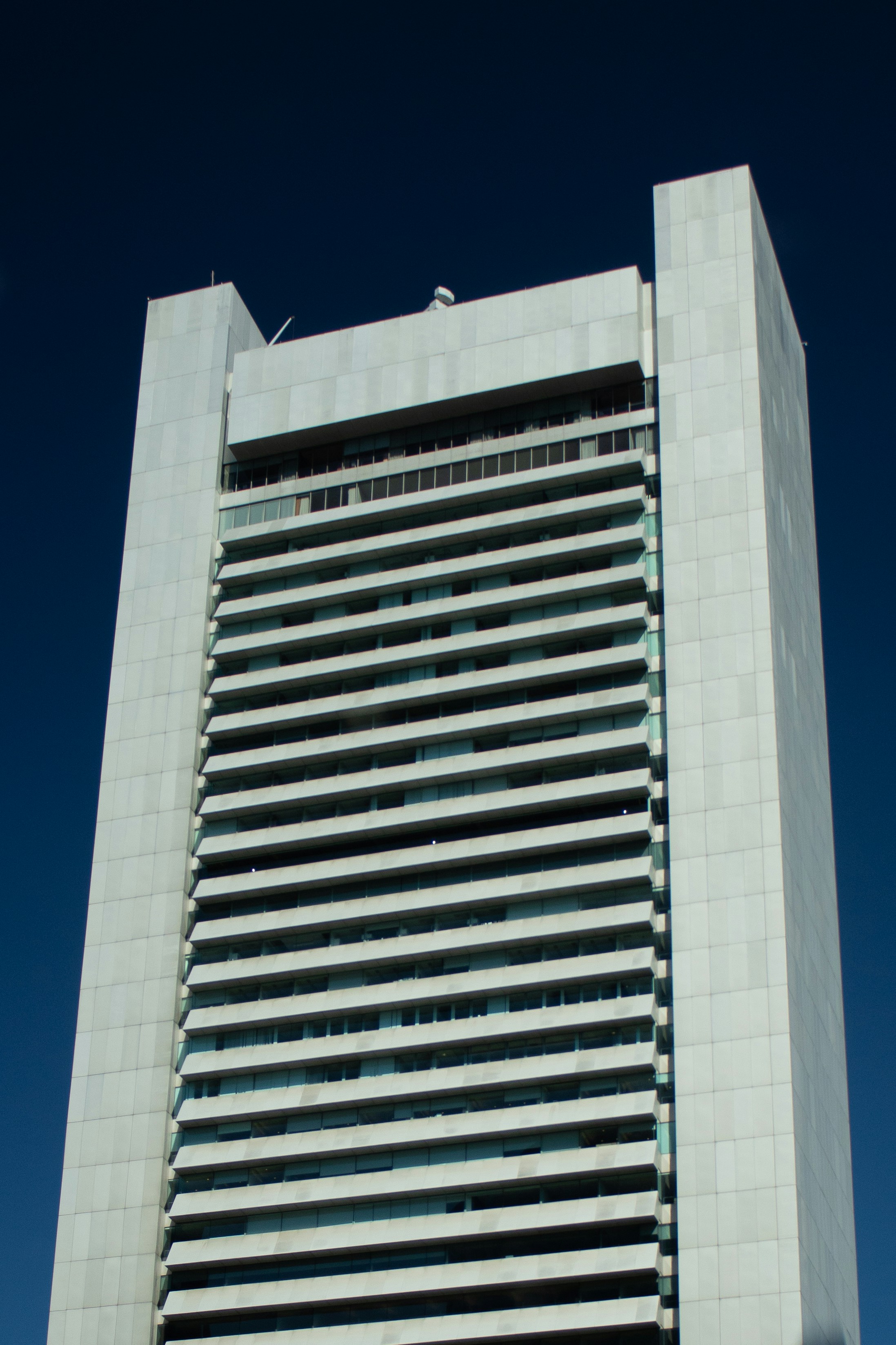 A striking high-rise building framed sharply against a rich blue sky, showcasing minimalist lines and brutalist influence. The uniform façade and bold horizontal slats make this architectural portrait both commanding and serene. | A tall building stands against a blue sky.