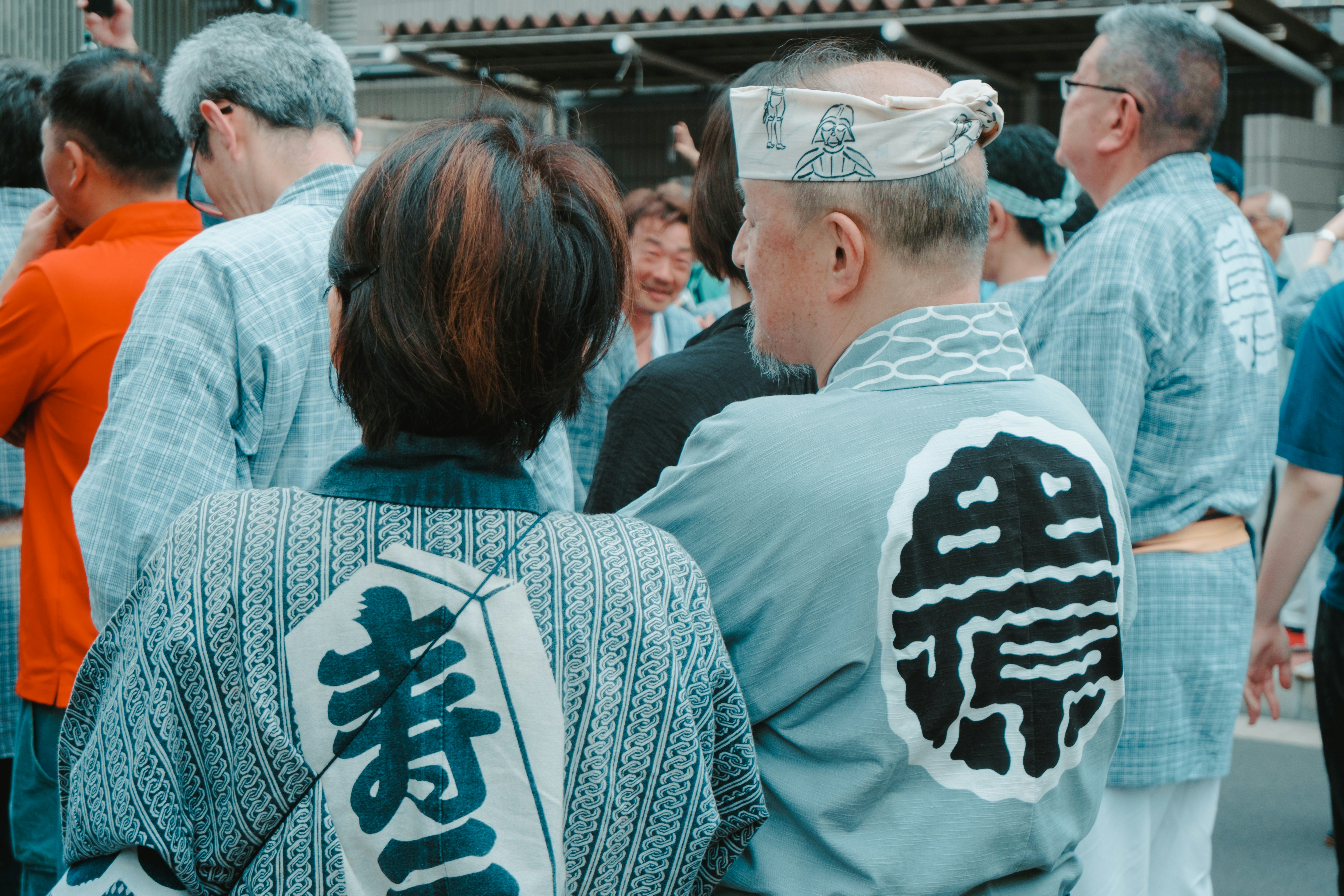 Two individuals in traditional attire stand amidst a crowd, showcasing intricate patterns on their garments. The scene captures the essence of a cultural celebration.