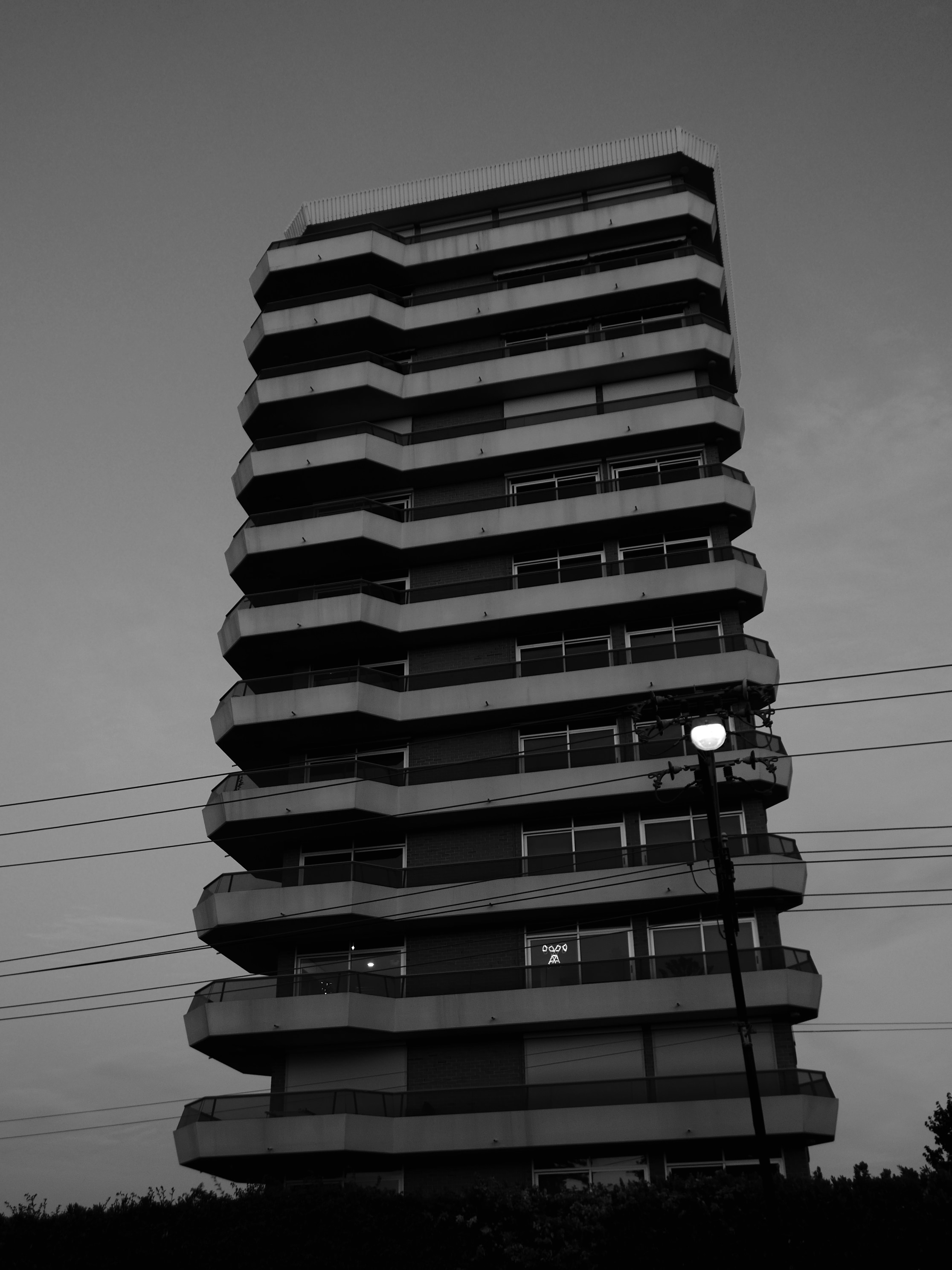 A sleek, modern building with staggered balconies against a twilight sky, showcasing geometric design. A solitary light illuminates a window, adding intrigue.