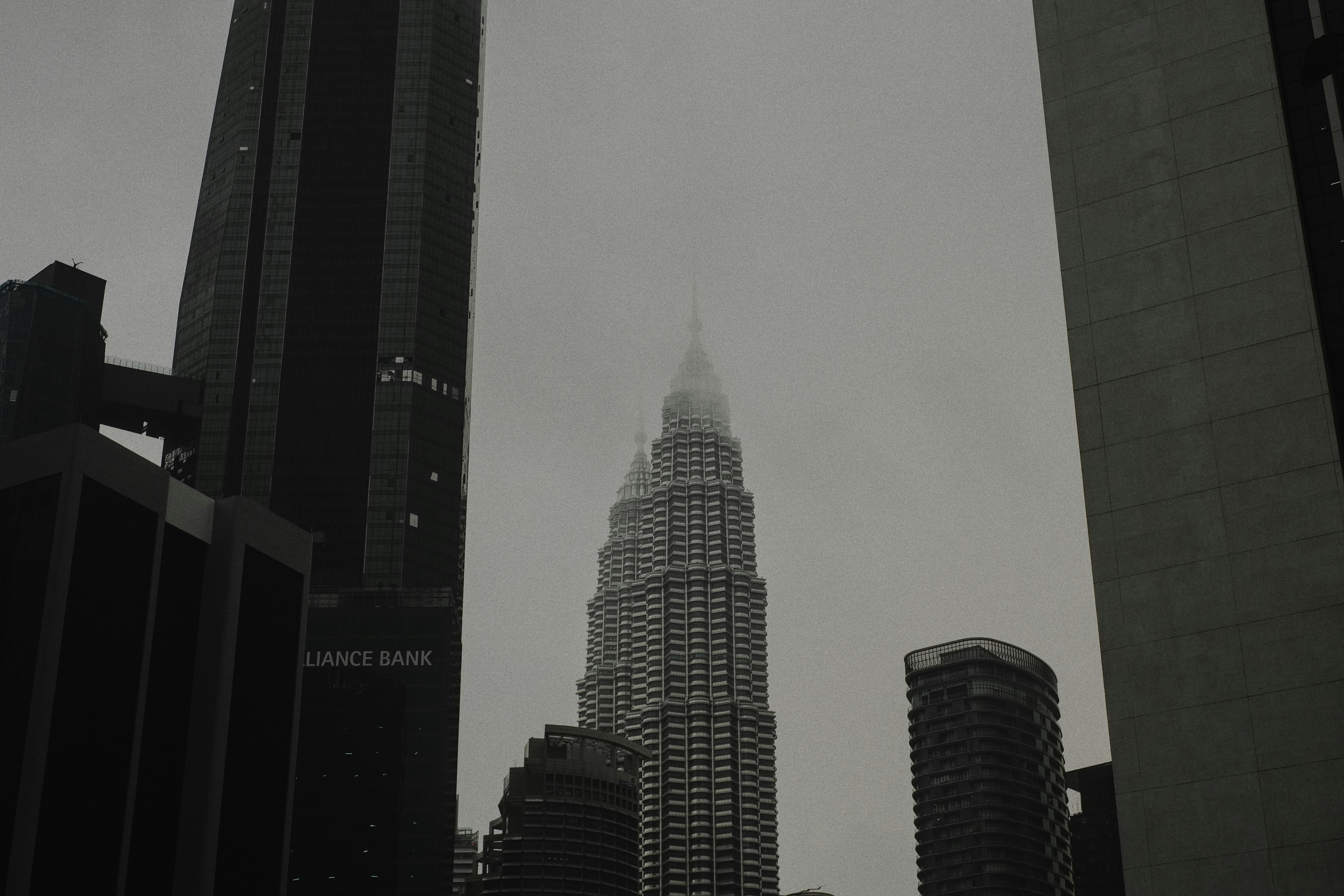 The iconic Petronas Towers rise through a veil of mist, flanked by modern skyscrapers in a monochromatic cityscape.