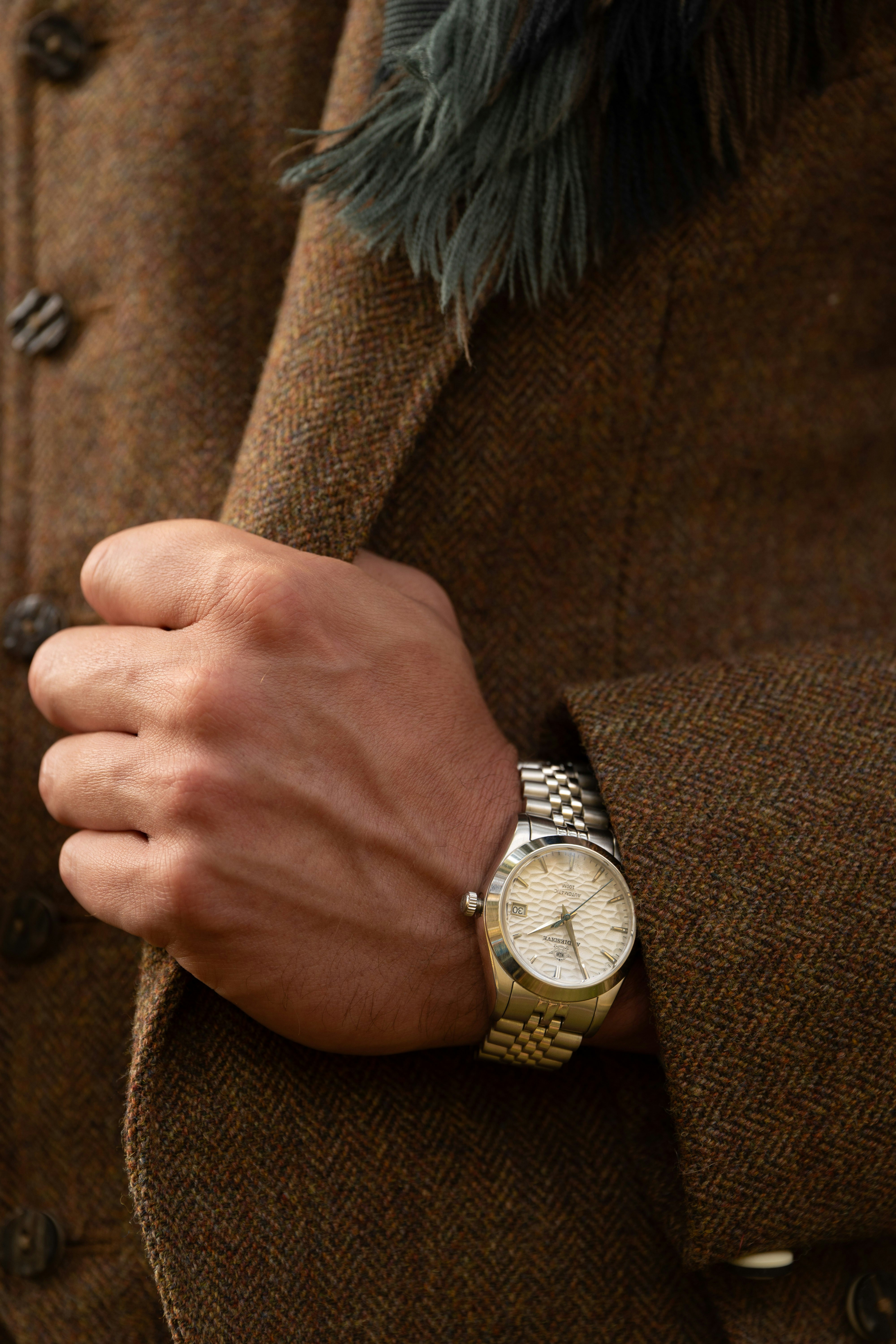 Close-up of a hand adjusting a coat, showcasing a classic wristwatch with a detailed dial. The textured fabric of the coat adds depth to the scene.