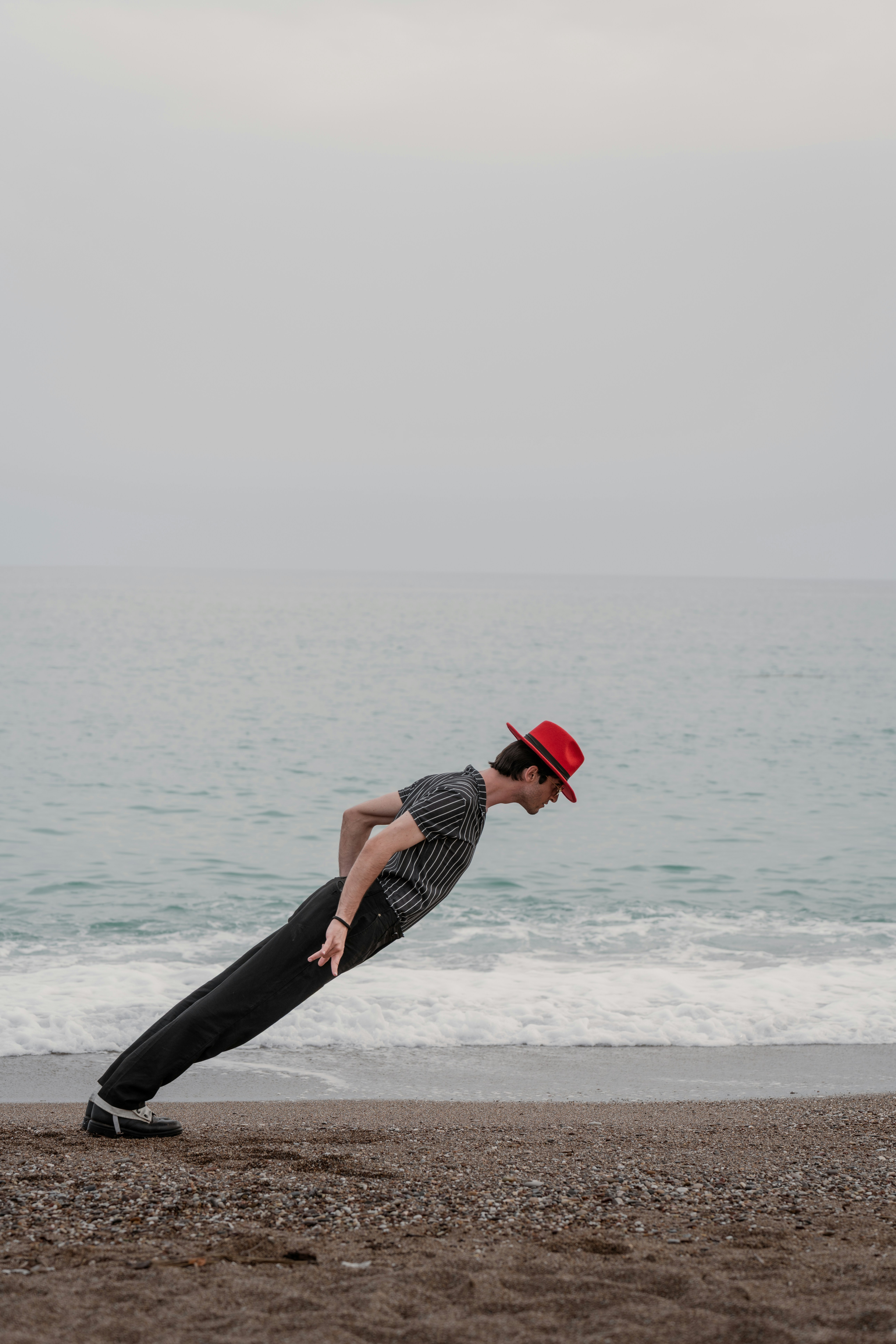 A man in a red hat leans dramatically over the shoreline, creating a playful visual illusion against a calm sea backdrop.