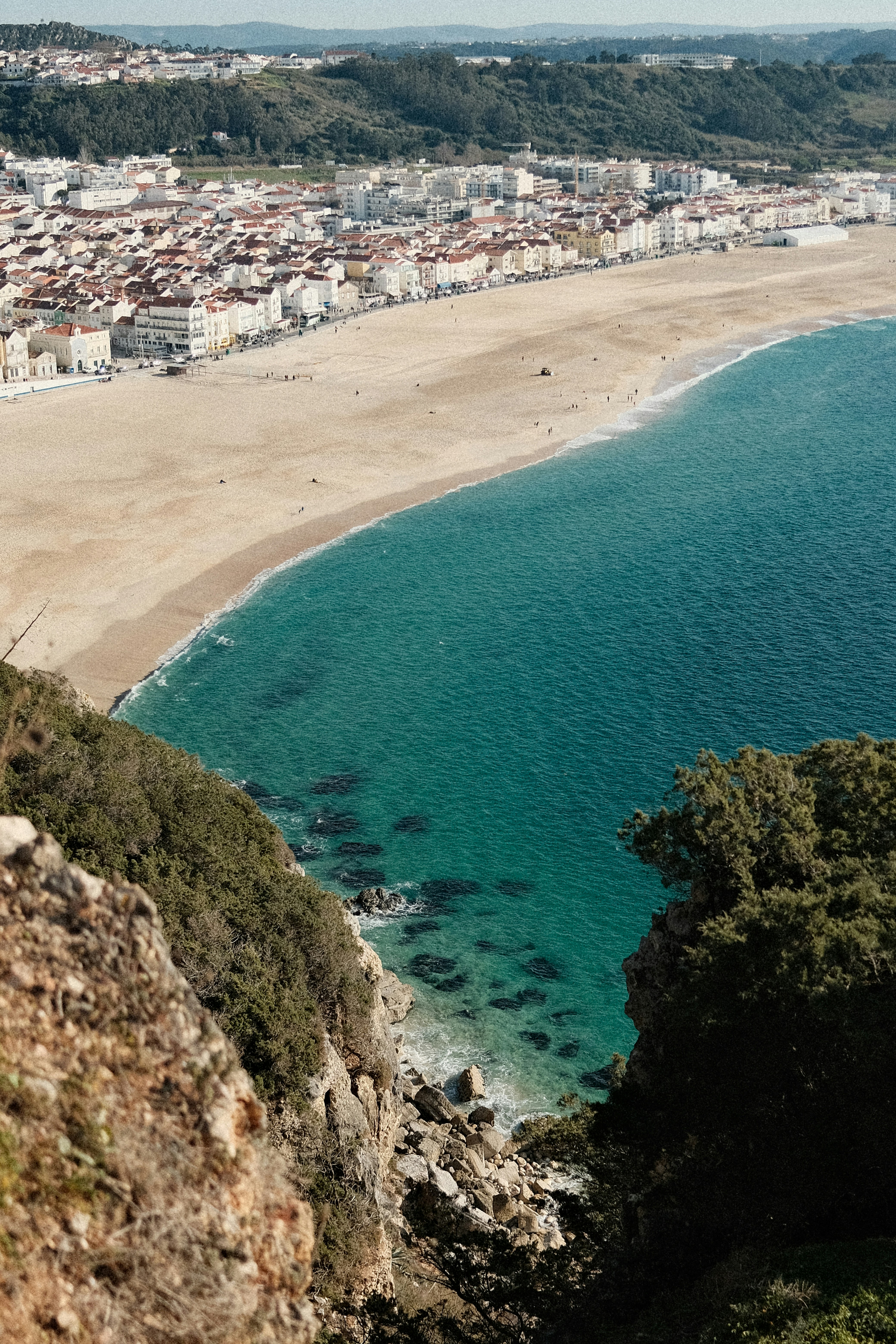 Aerial view of a sandy beach meeting a vibrant turquoise sea, bordered by lush greenery and a charming coastal town in the background.