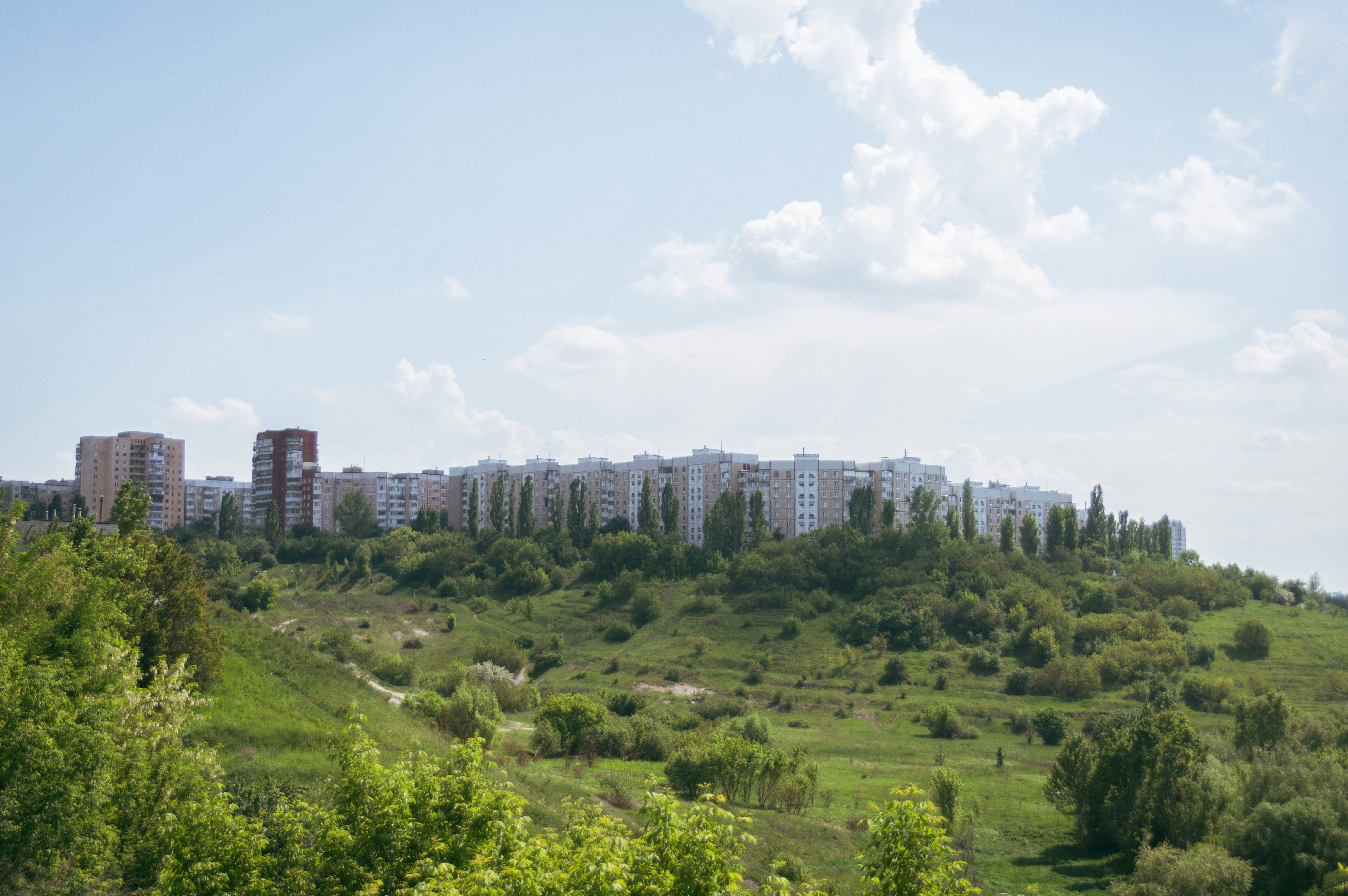 A general view of a natural landscape with hills and multi-story buildings in the distance in summer. | Distant buildings sit atop a green hillside.