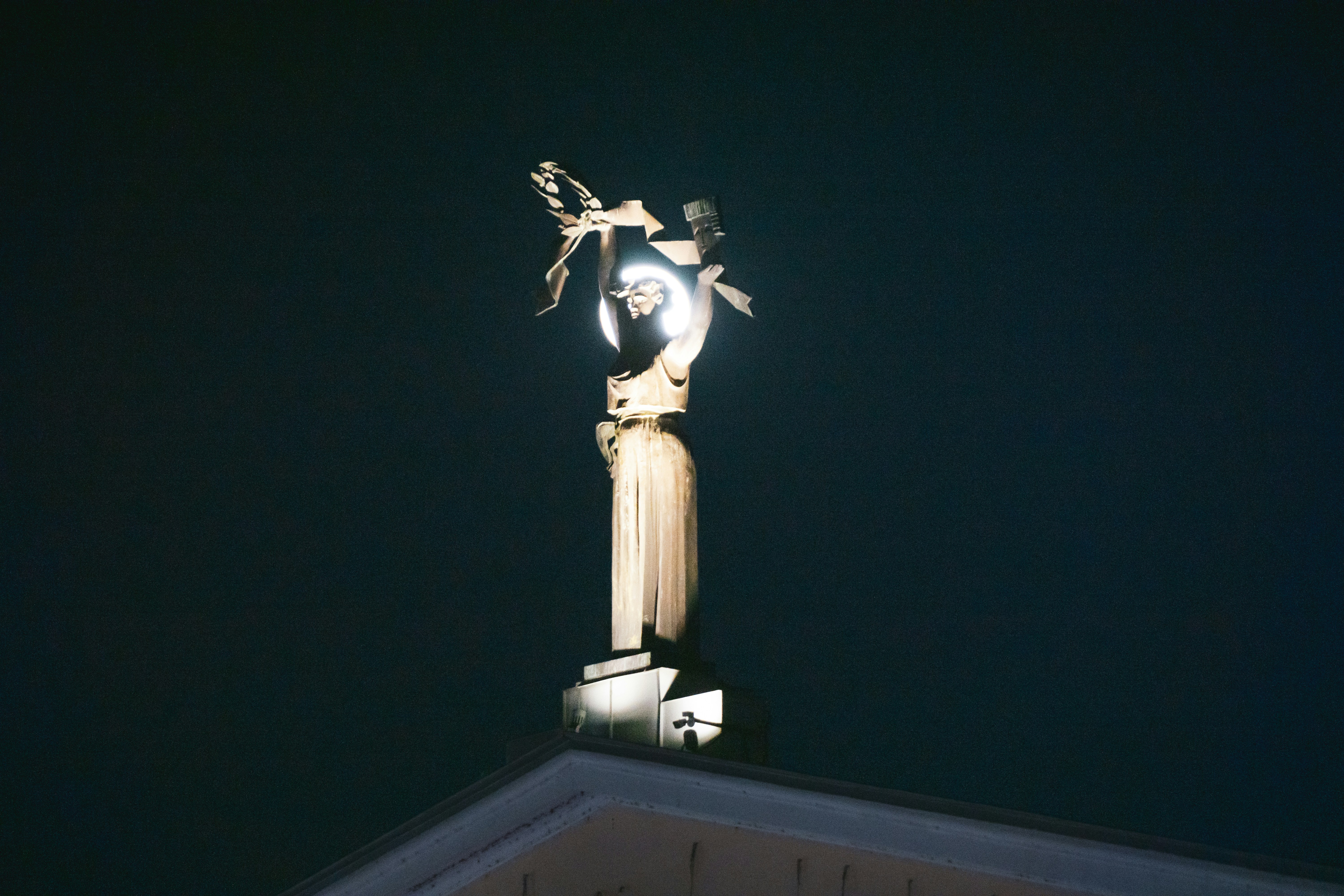 A luminous statue holding a laurel wreath and a sword, illuminated against a dark sky. The figure stands atop a building, symbolizing victory and peace.