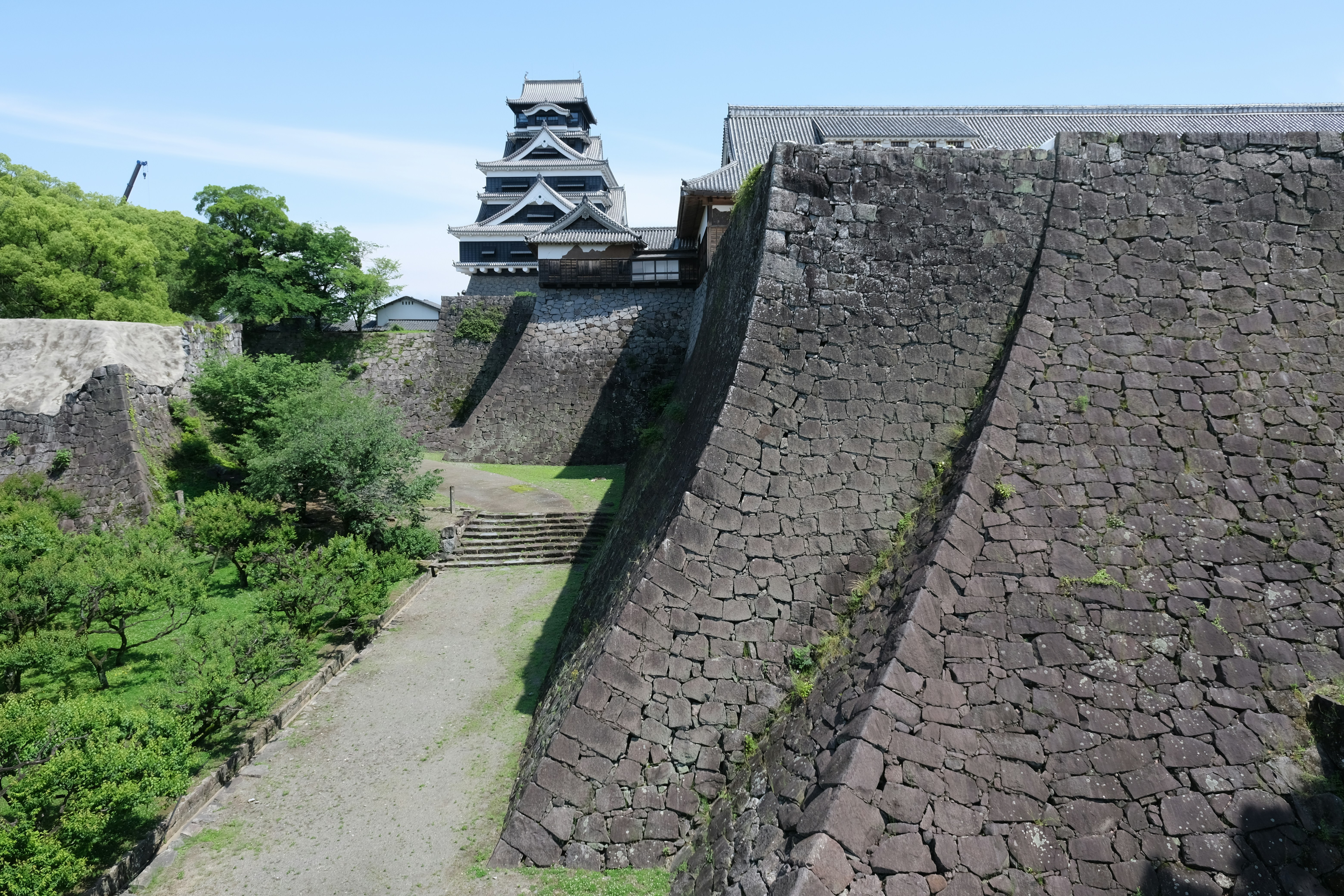 Kumamoto Castle's imposing stone walls and traditional architecture stand against a clear blue sky, surrounded by lush greenery. The pathway leading up to the castle invites exploration.