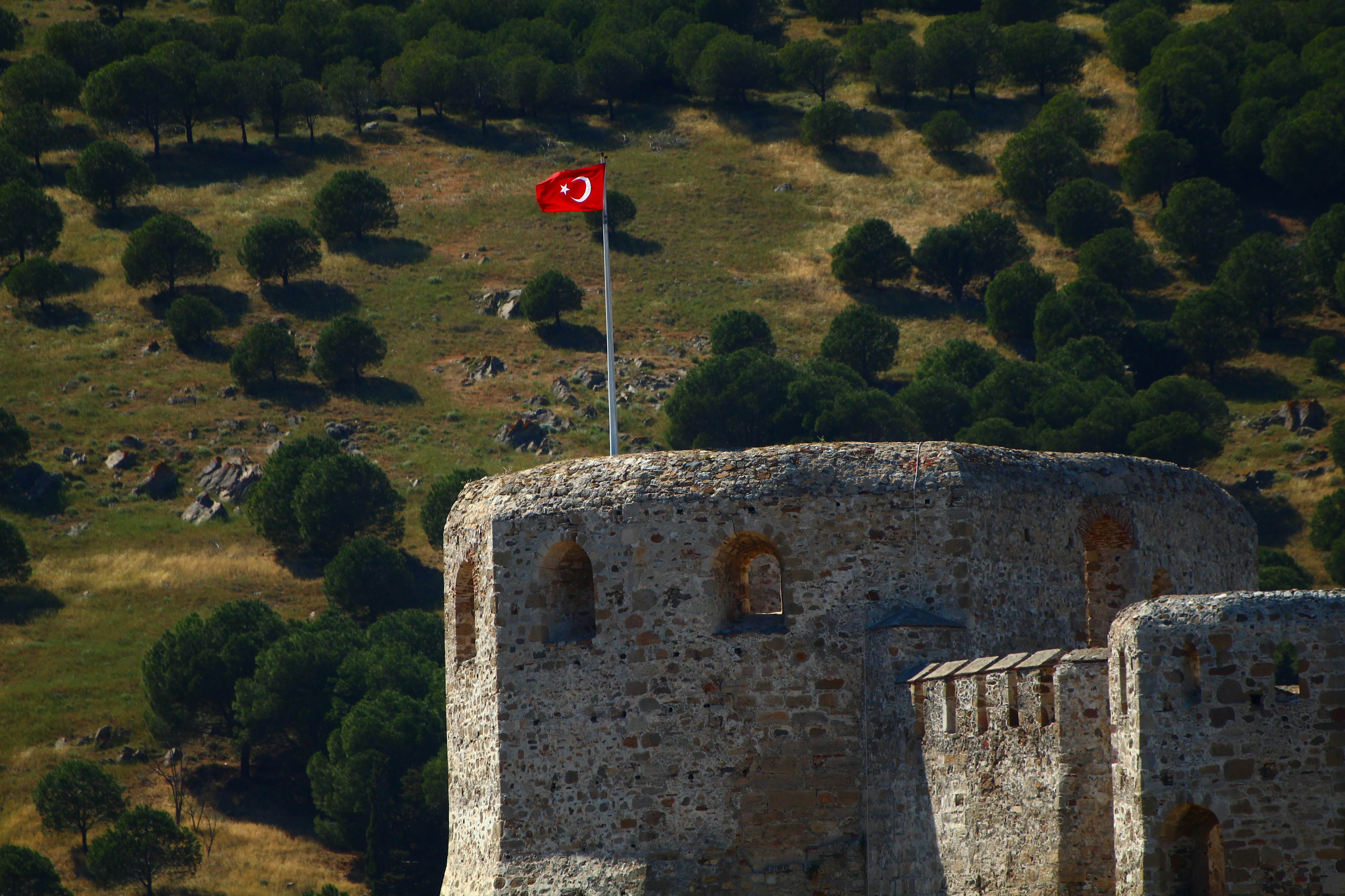 Historic stone fortress prominently displaying the Turkish flag against a backdrop of rolling green hills.