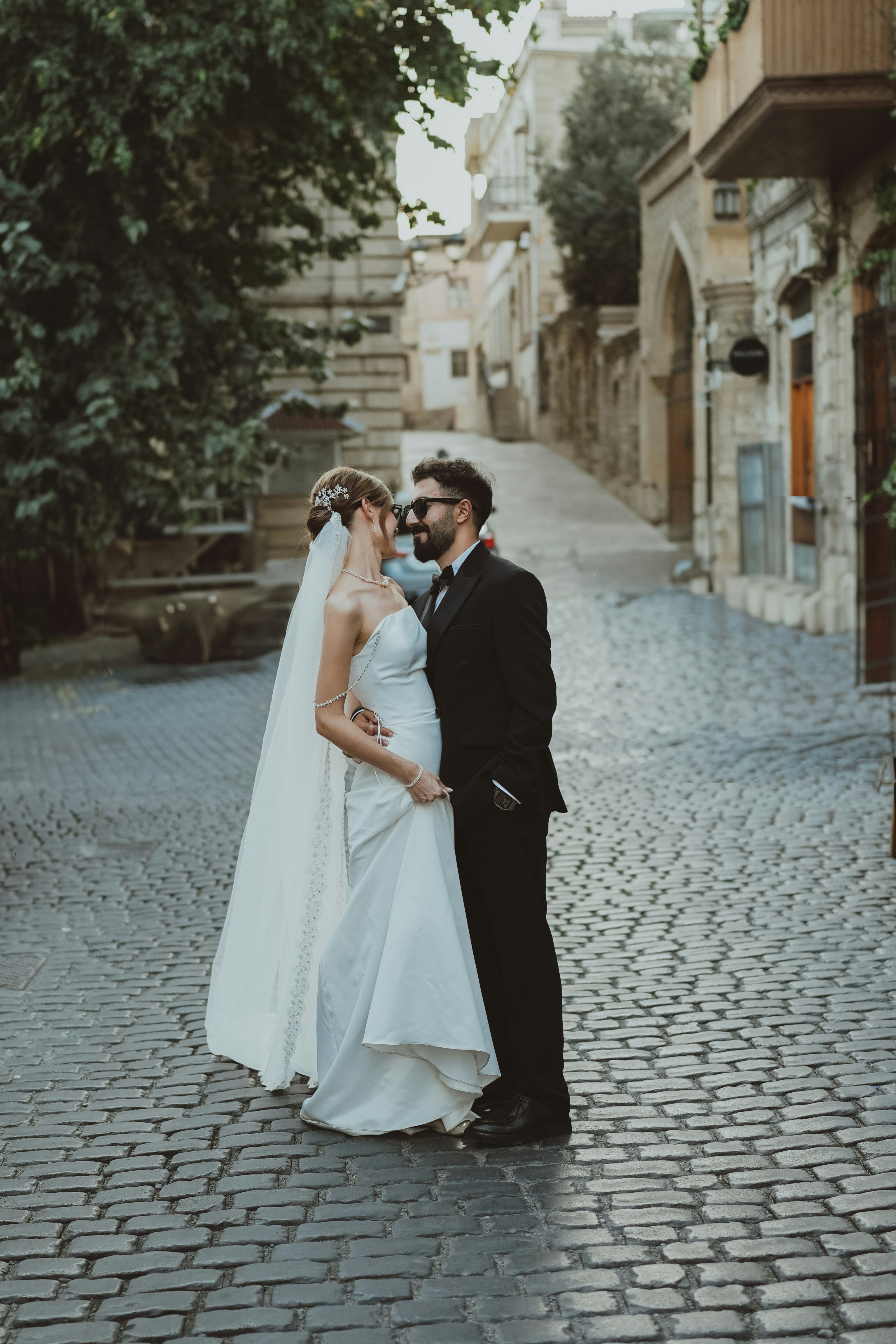 Bride and groom sharing a tender moment on a cobblestone street, surrounded by historic architecture and lush greenery.