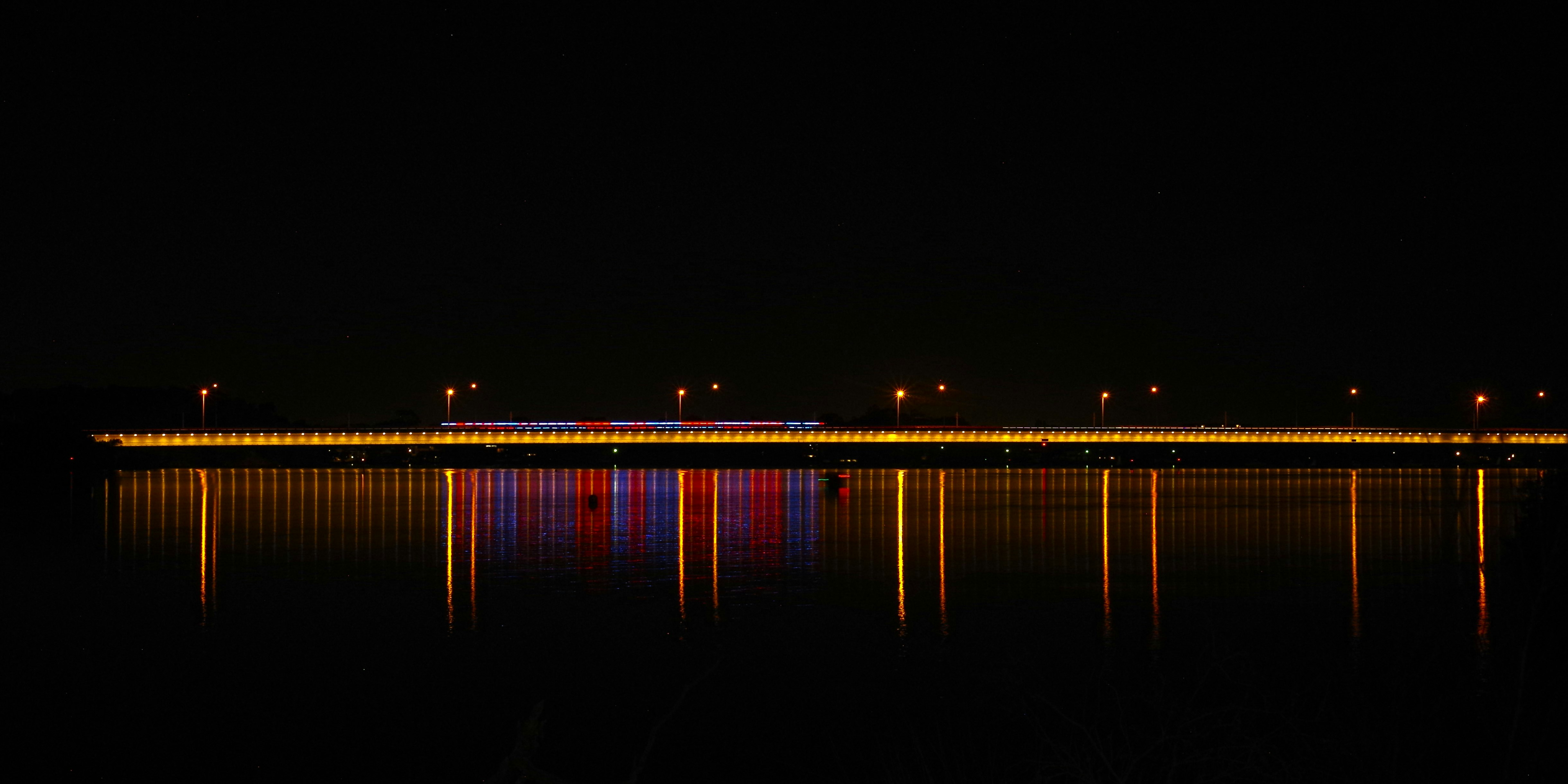 A brightly lit bridge spans across a calm body of water, with vibrant reflections creating a mesmerizing visual effect.