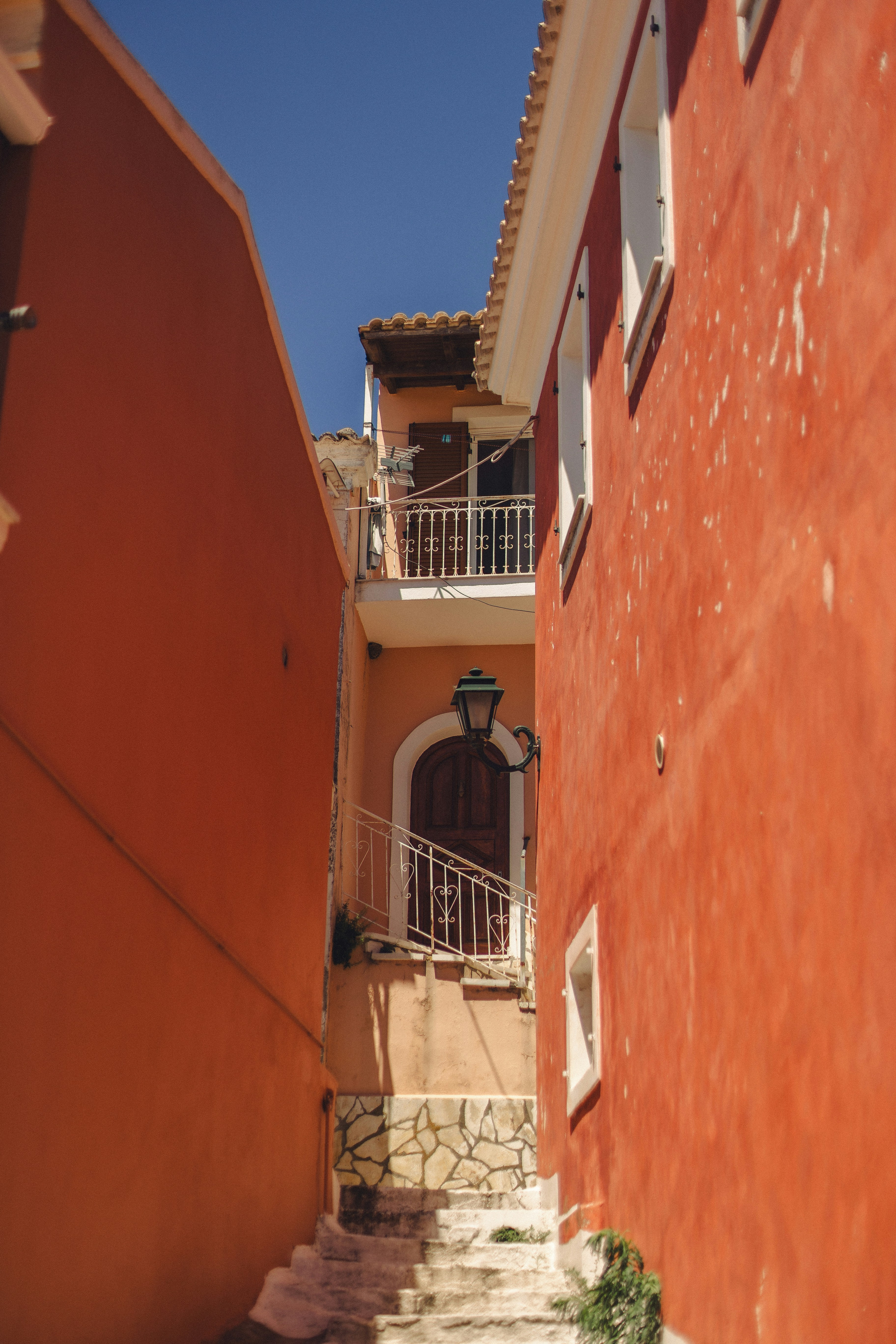 Narrow alleyway flanked by vibrant orange walls leading to a wooden door with a balcony above. A vintage lantern adds character to the scene.