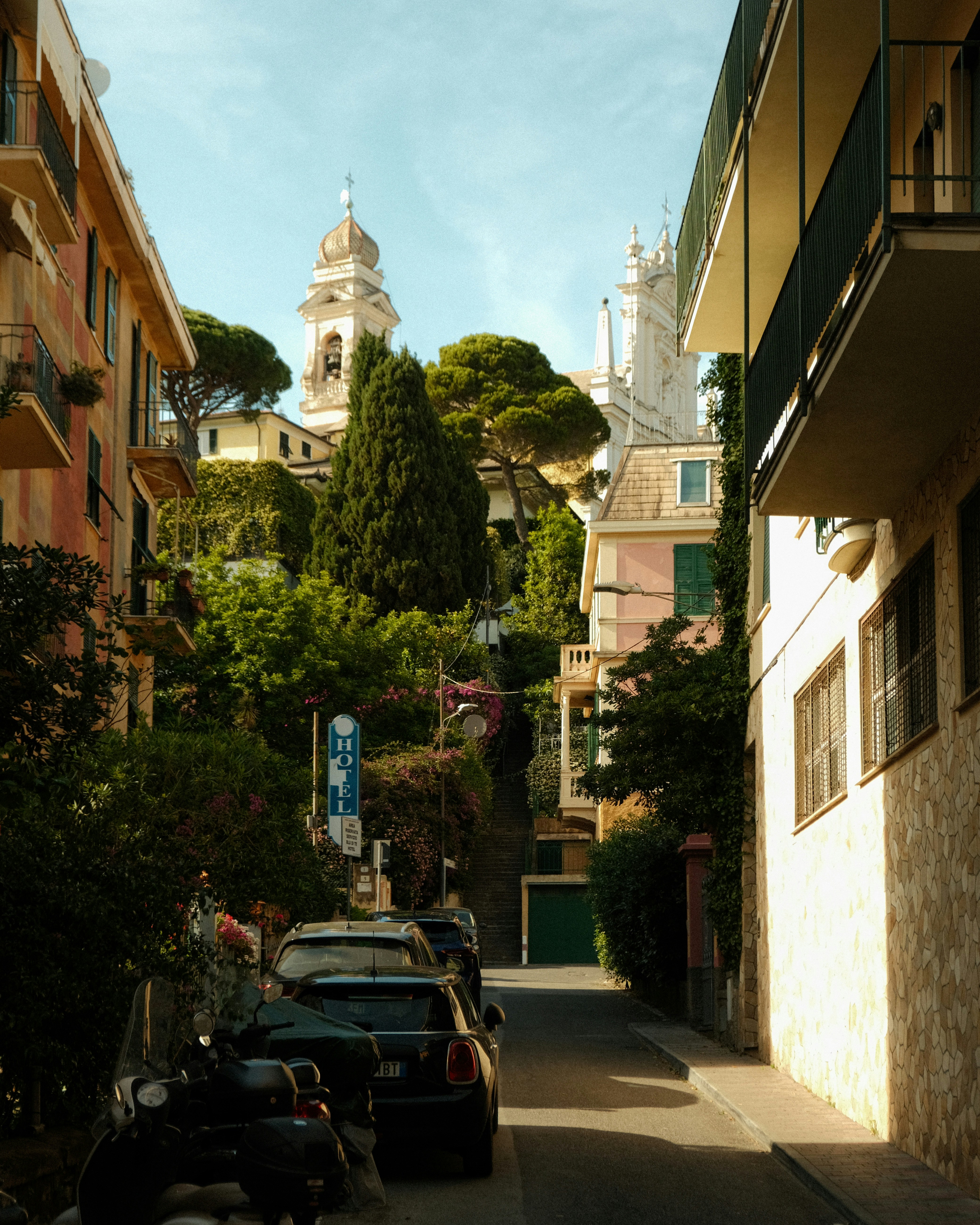 Narrow street lined with colorful buildings and lush greenery leading up to a church tower, capturing the essence of a quaint coastal town.