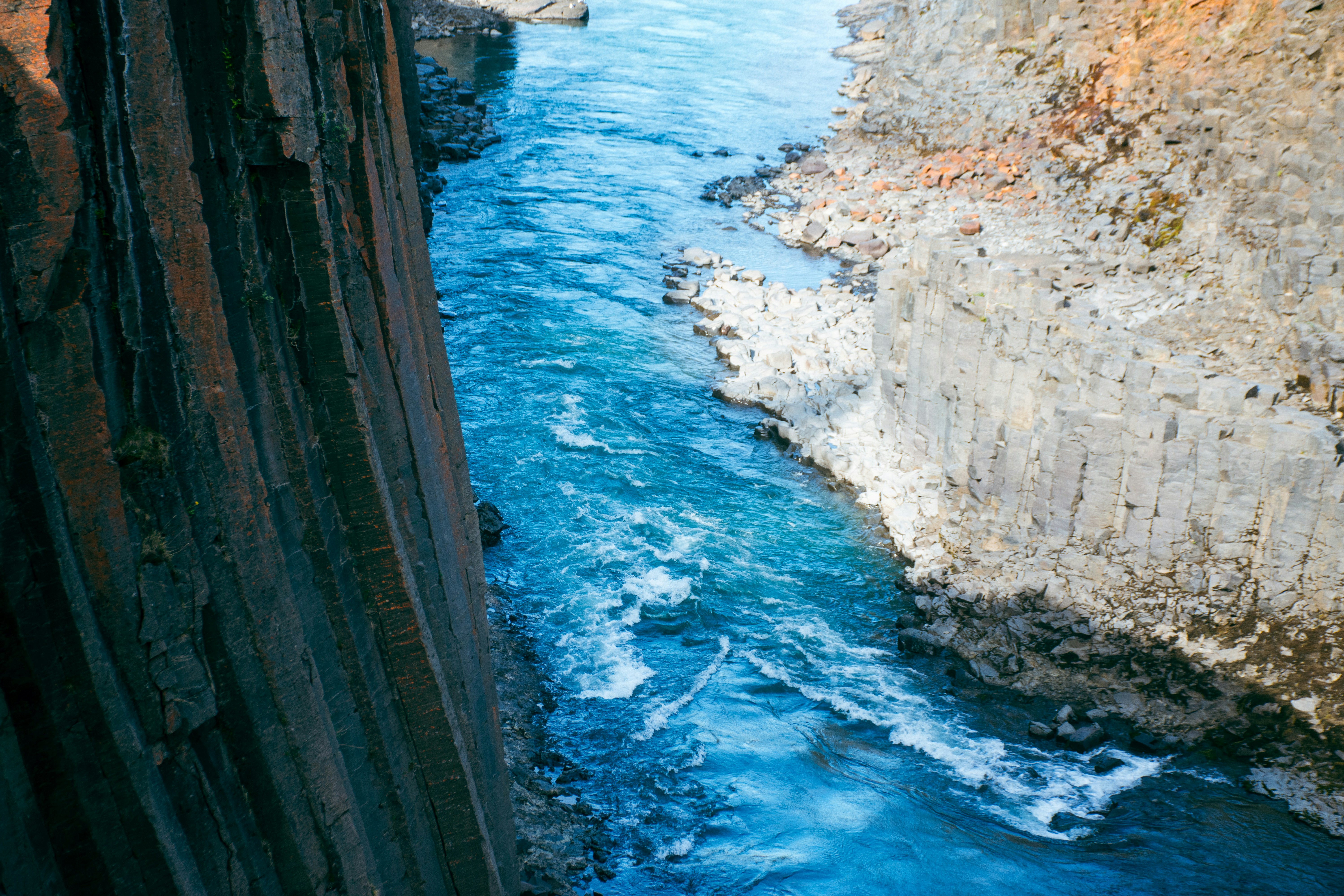 Turquoise waters meander between towering basalt cliffs, showcasing the intricate interplay of nature's forces. The scene captures the raw beauty of the landscape.