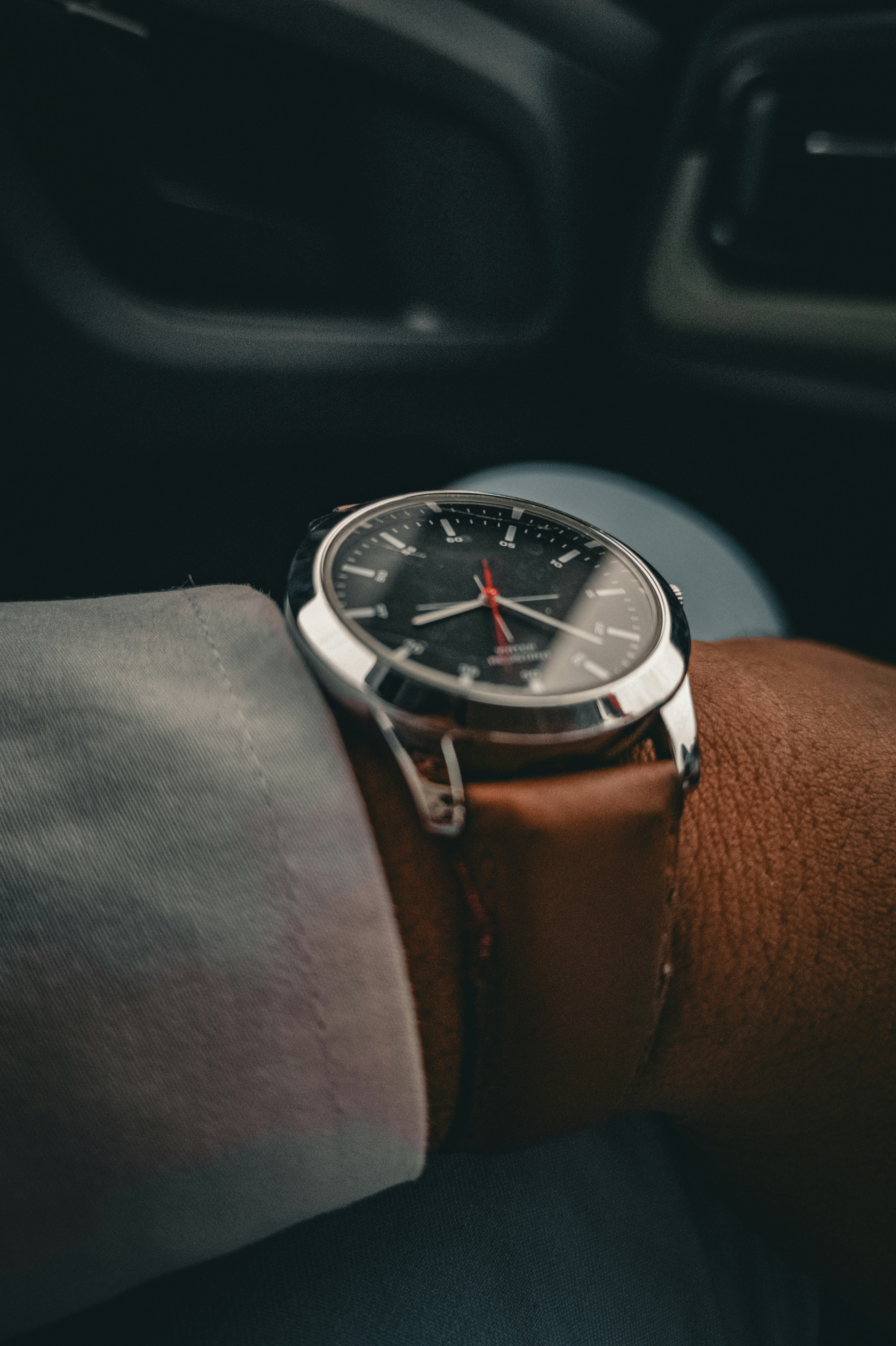 The watch has a black face with silver hands and a red second hand. The image is taken from a slightly angled perspective, giving a sense of depth and focus on the watch. The background is blurred, which helps to draw attention to the watch and the wrist. | A wristwatch worn by someone in a car.