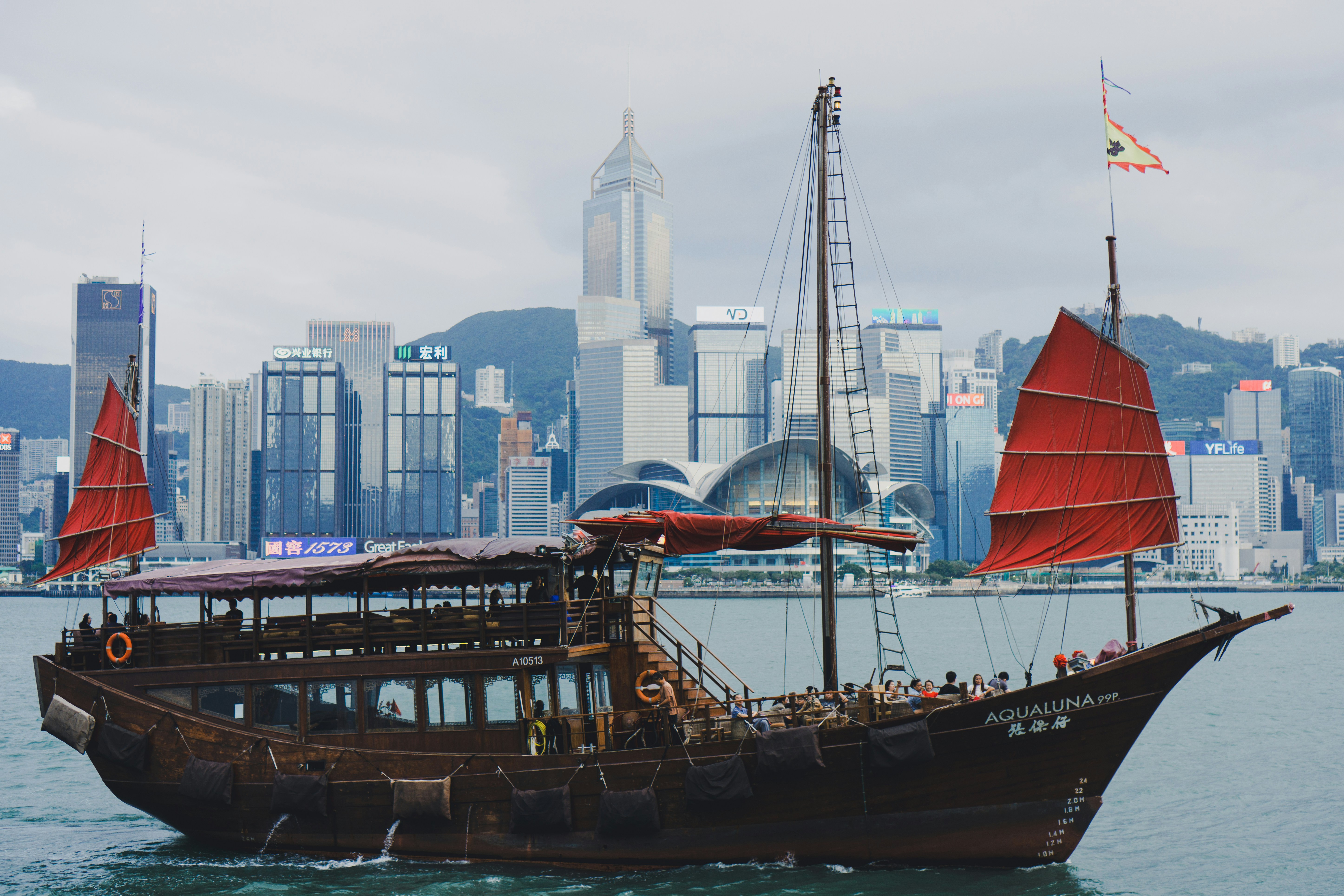 The distinctive red sails of the Aqua Luna, a handcrafted Chinese junk, cut a striking silhouette against the modern Hong Kong skyline across Victoria Harbour. Its dark, traditional wooden hull, meticulously crafted by an 80-year-old local artisan, carries passengers enjoying the scenic journey. Named "Cheung Po Tsai" in Chinese after a legendary pirate, the vessel evokes a rich maritime history, contrasting sharply with the gleaming skyscrapers and urban sprawl in the background. | A chinese junk boat sails in front of a city.