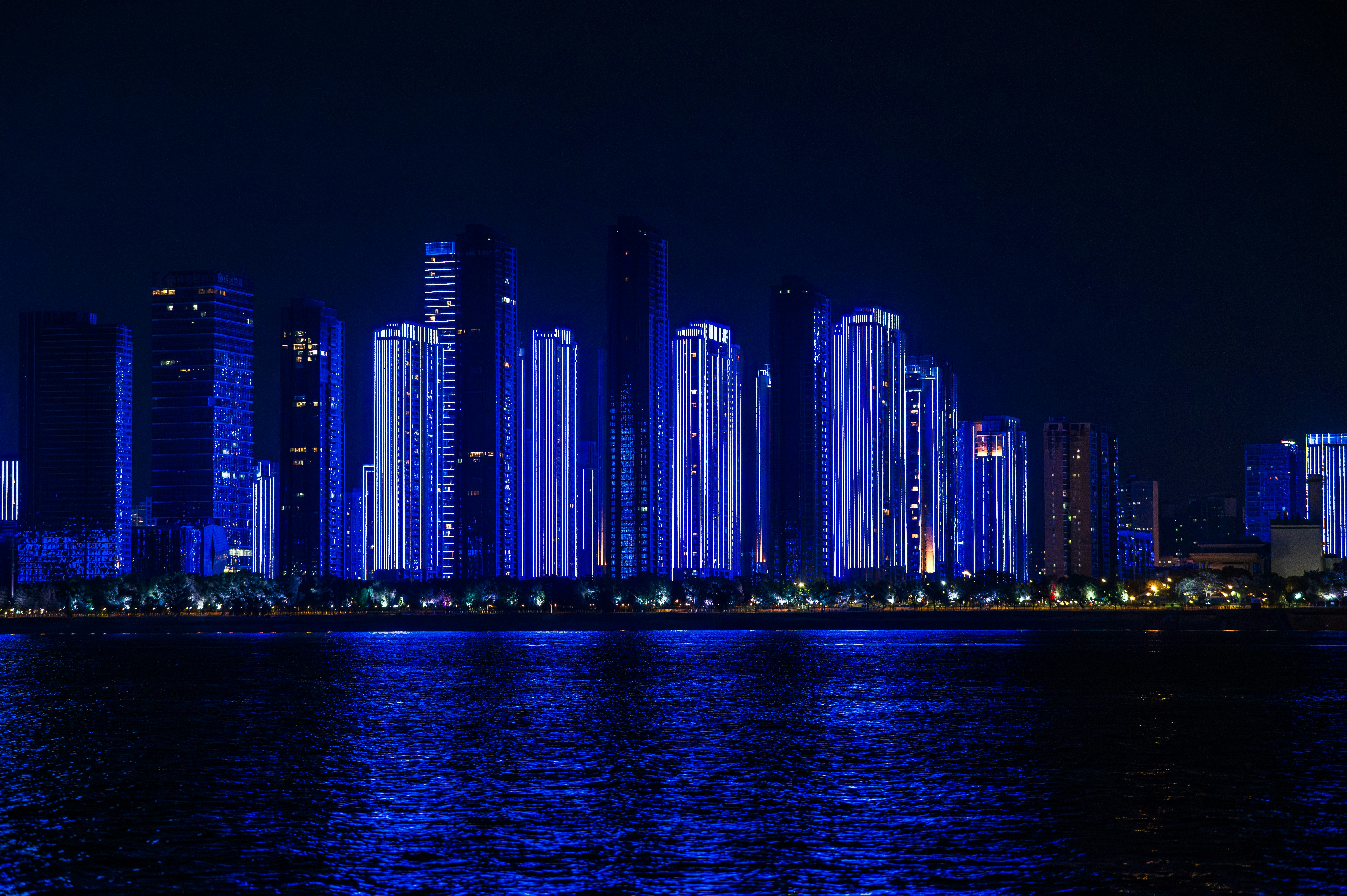 Illuminated skyscrapers reflecting on water under a night sky, showcasing vibrant blue lights and urban architecture.