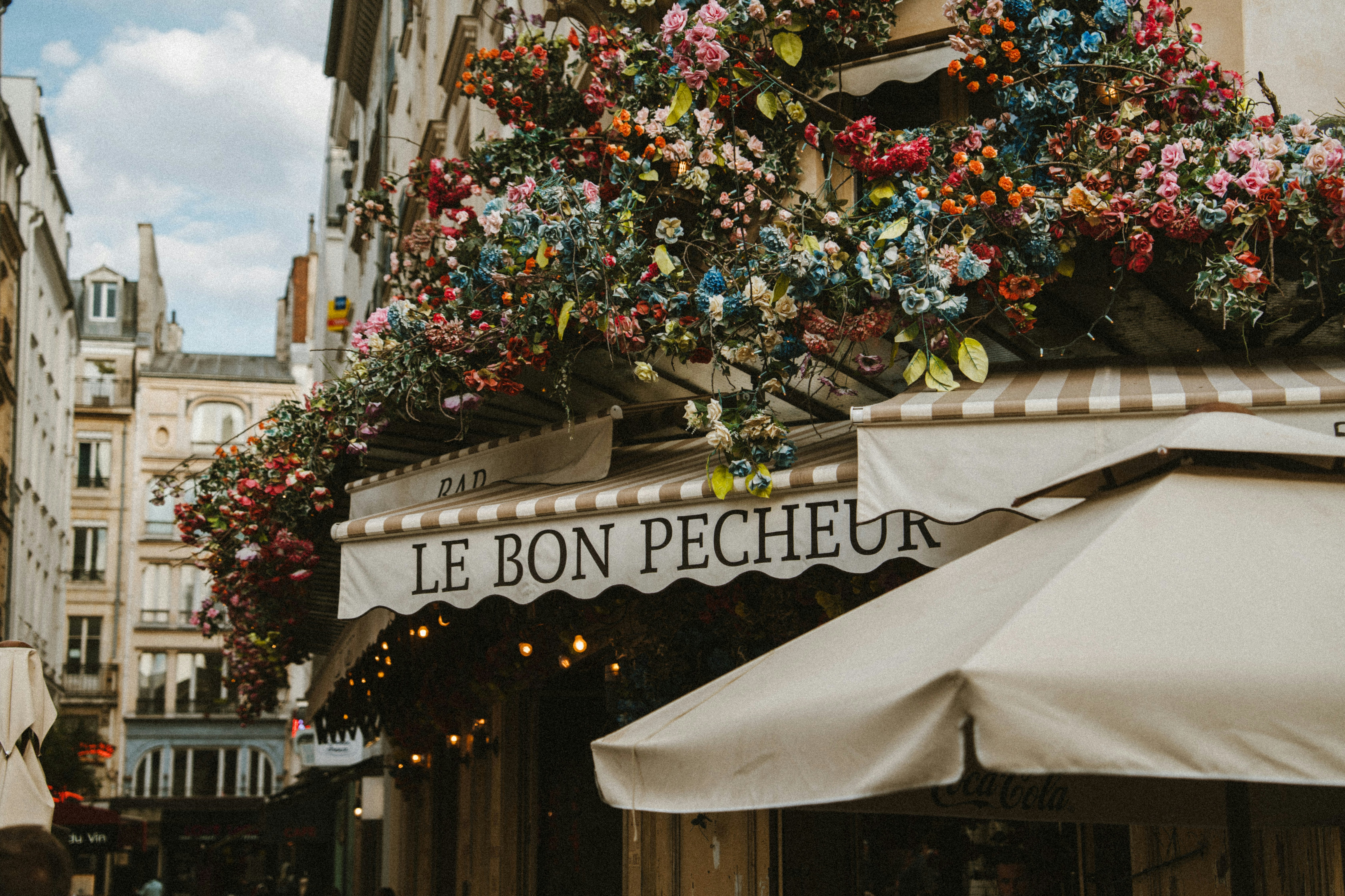 Vibrant flowers cascade over a quaint Parisian restaurant awning, inviting passersby to enjoy the ambiance. The establishment's name, 'LE BON PECHEUR,' is elegantly displayed.