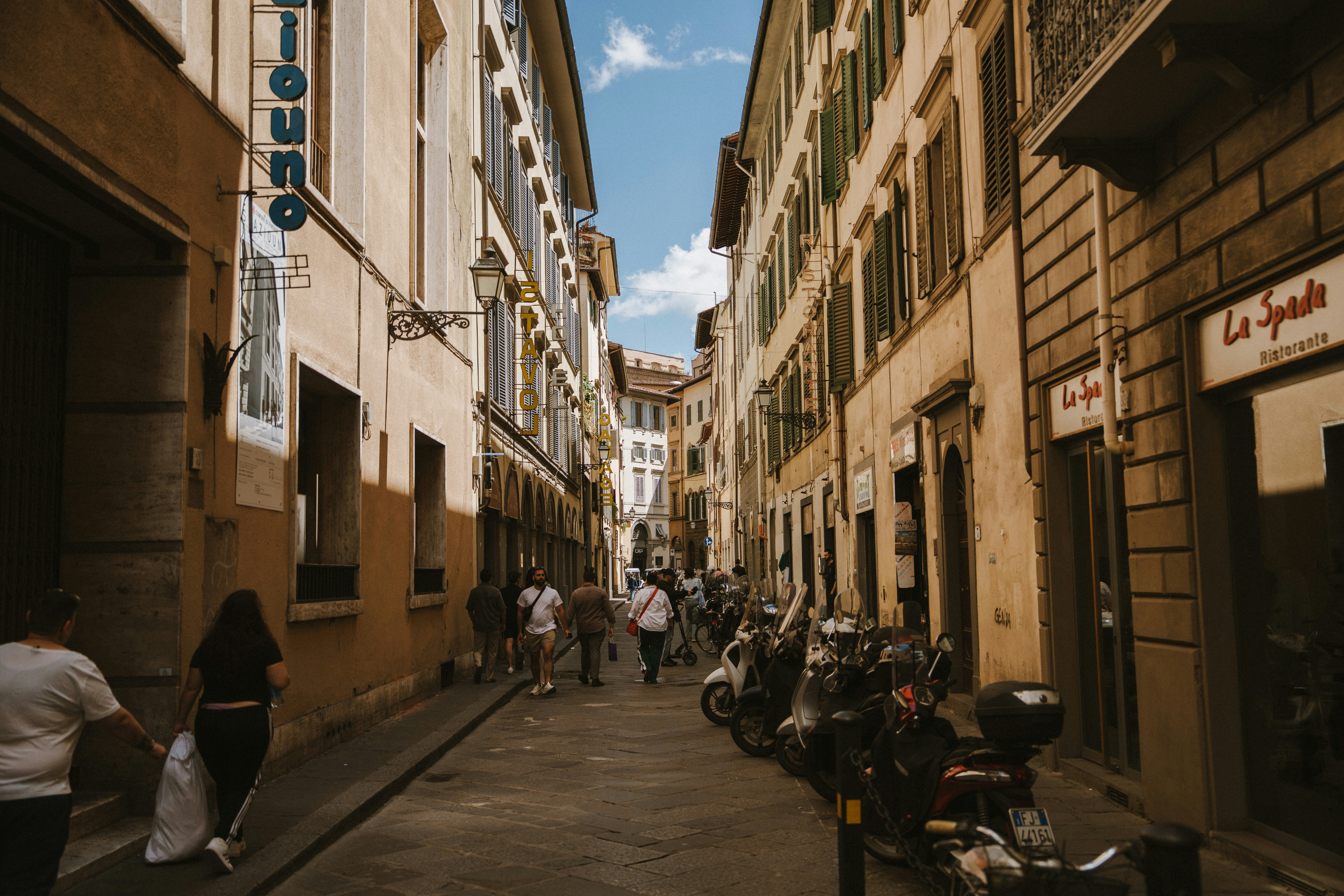 Charming narrow street lined with historic buildings, scooters parked along the side, and pedestrians enjoying the day.