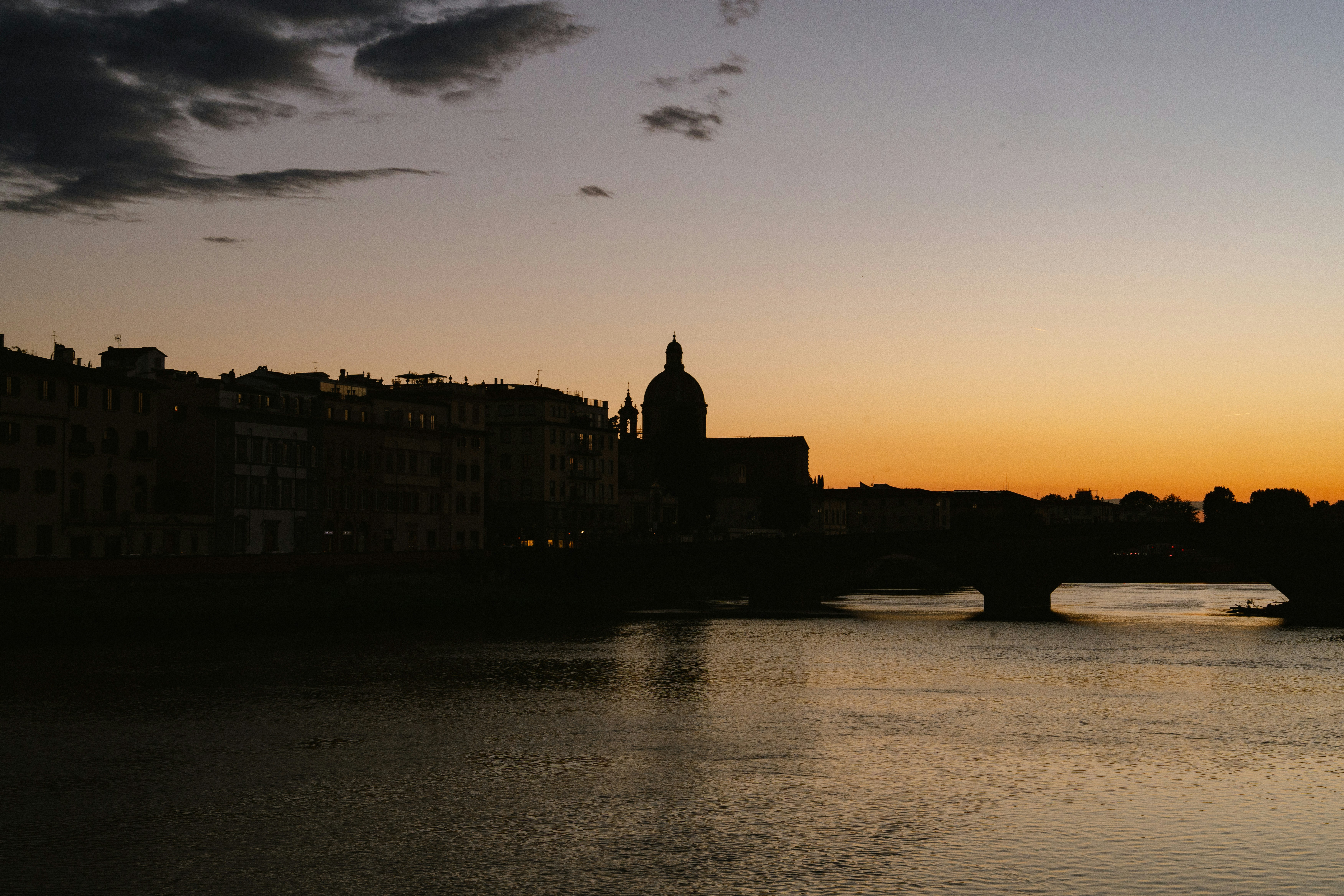 Silhouetted cityscape along the Arno River at dusk, highlighting historical architecture against a gradient sky. The scene captures the tranquil essence of evening in Florence.