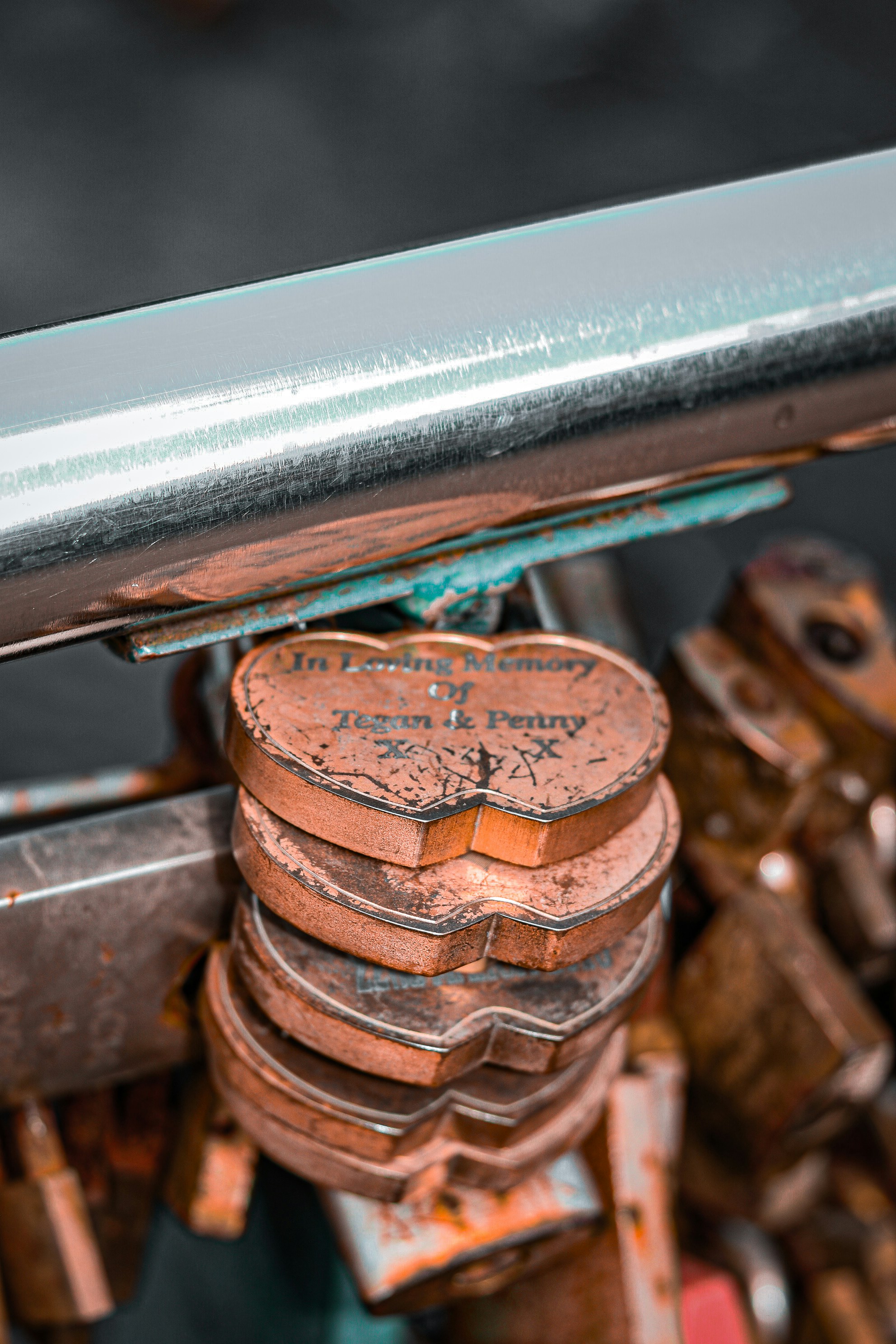 Stack of heart-shaped locks inscribed with a memorial message, symbolizing love and remembrance. The locks are attached to a metallic railing, showcasing a blend of rust and shine.