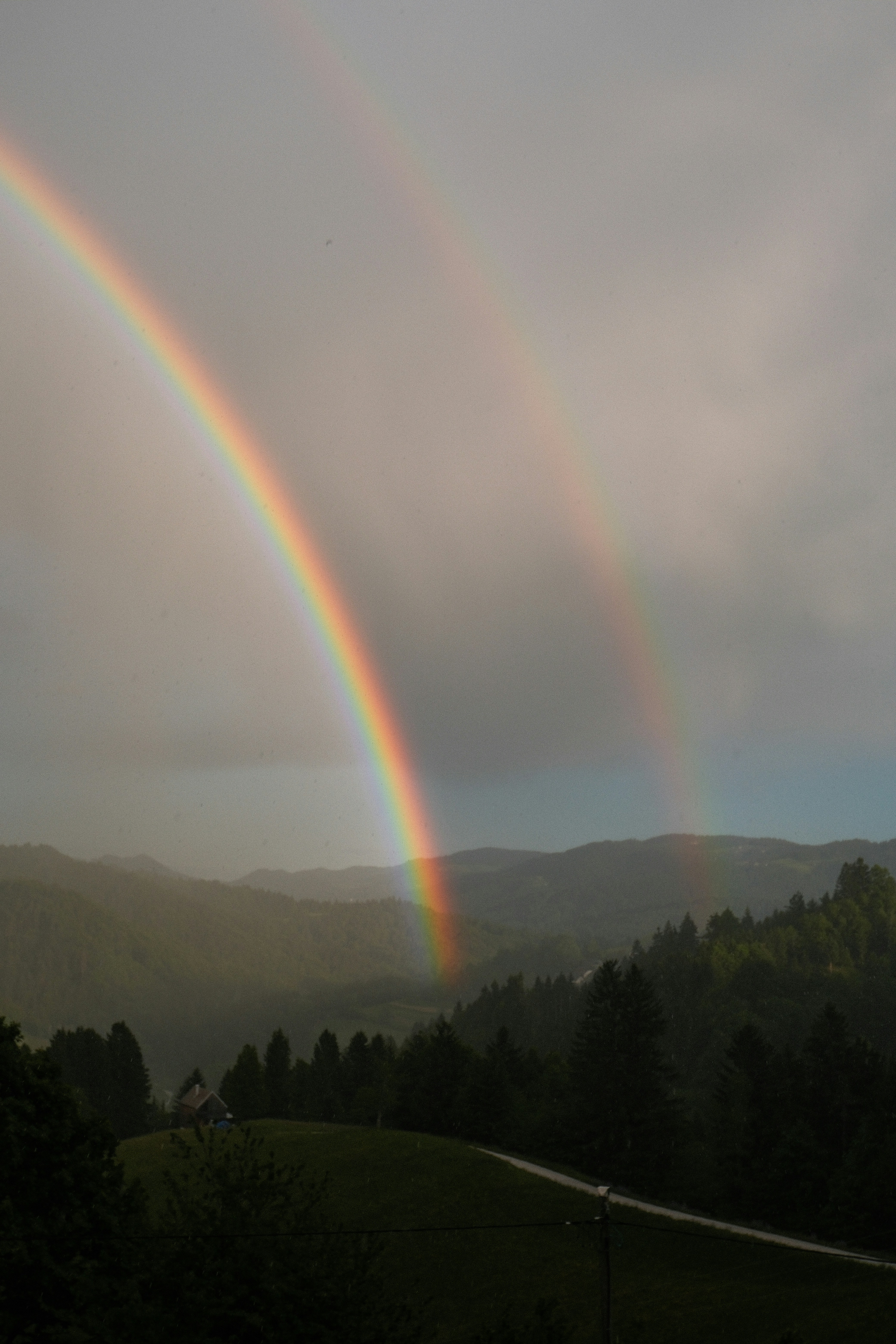 A vibrant double rainbow arcs over a lush mountain landscape, framed by dark clouds and a serene countryside. The scene captures the fleeting beauty of nature's spectrum.